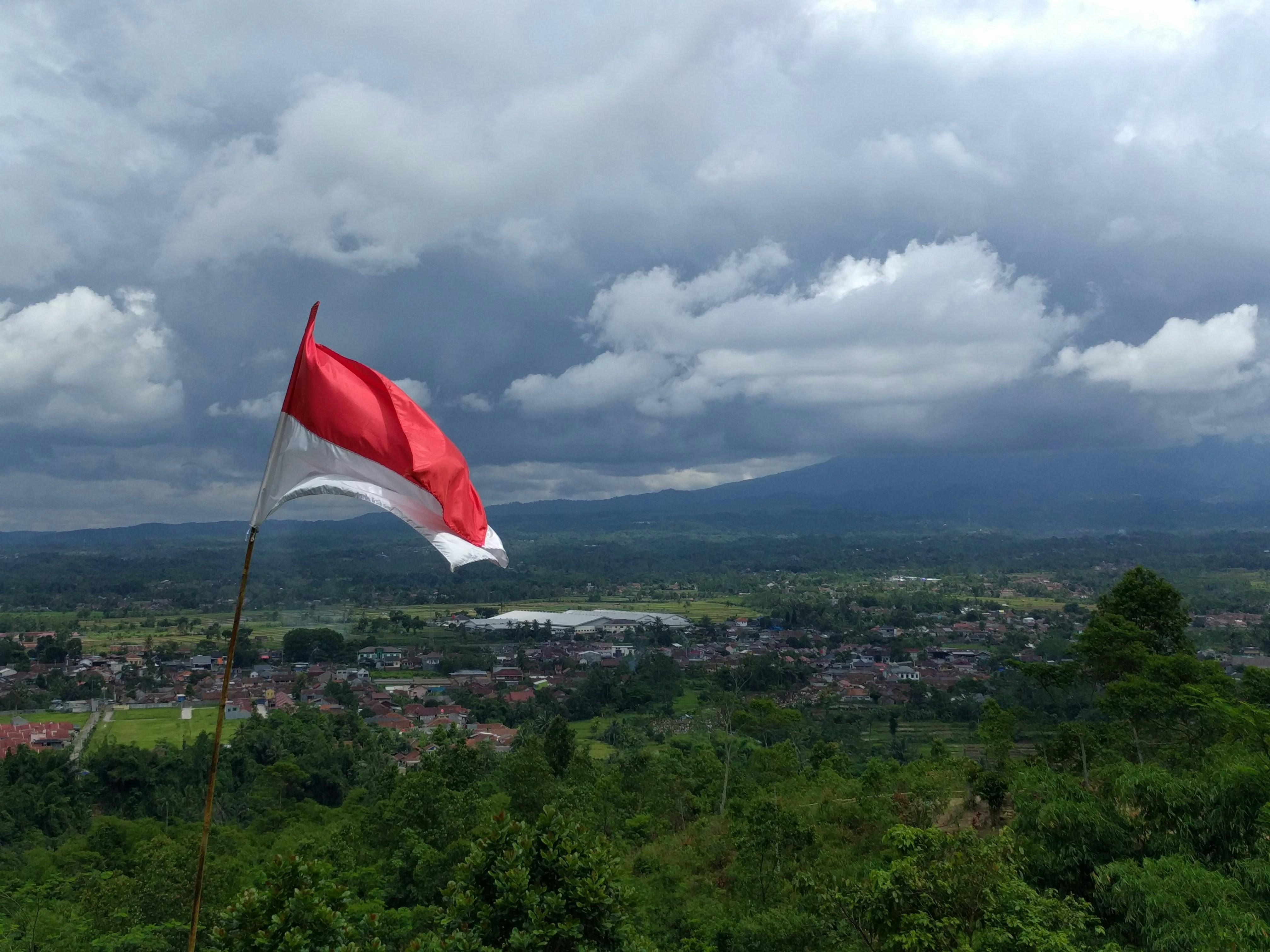 Indonesian flag waves over lush landscape with distant mountains under a cloudy sky.