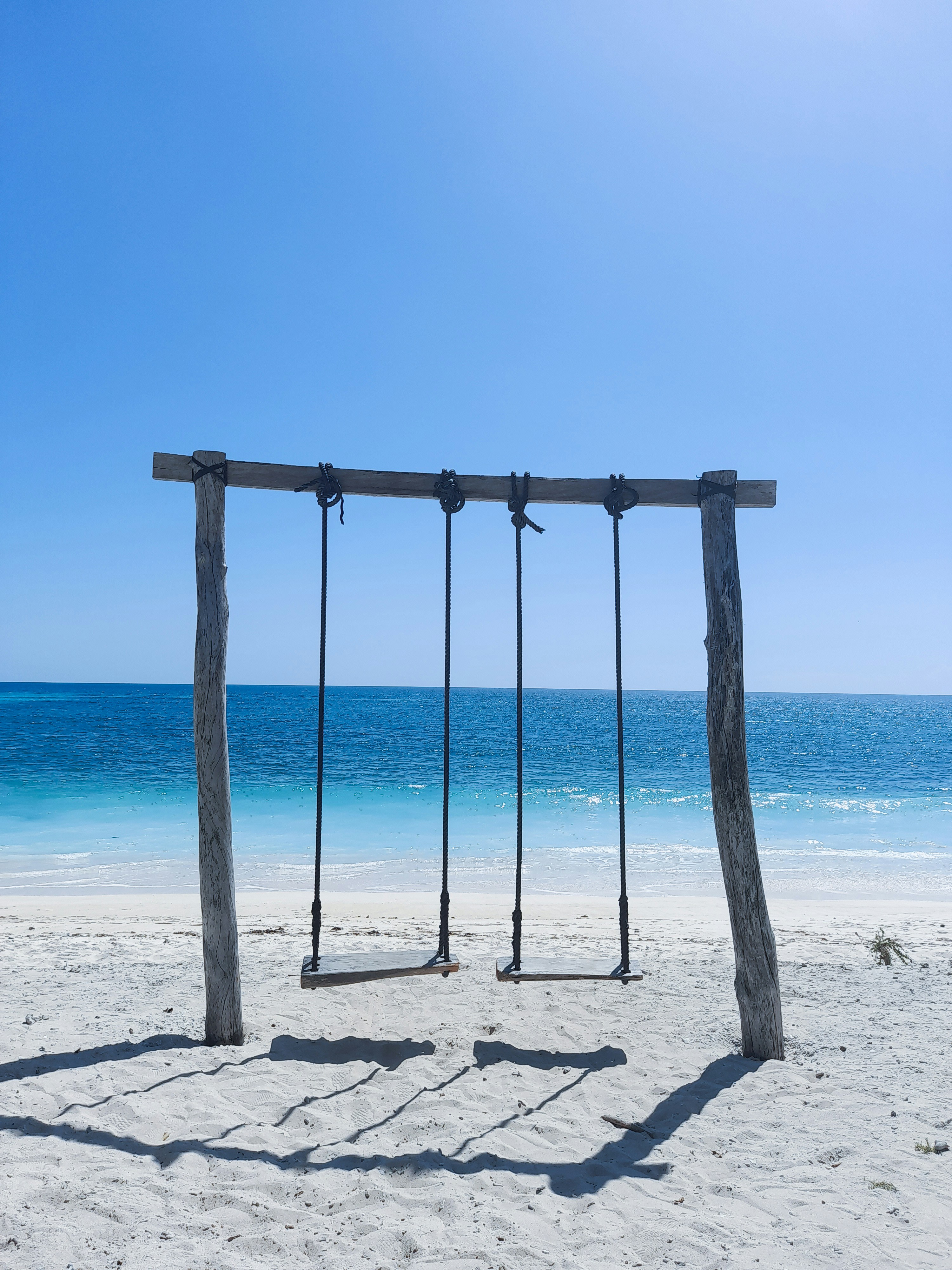 Wooden swings stand on a pristine white sandy beach against a vibrant blue ocean under a clear sky.
