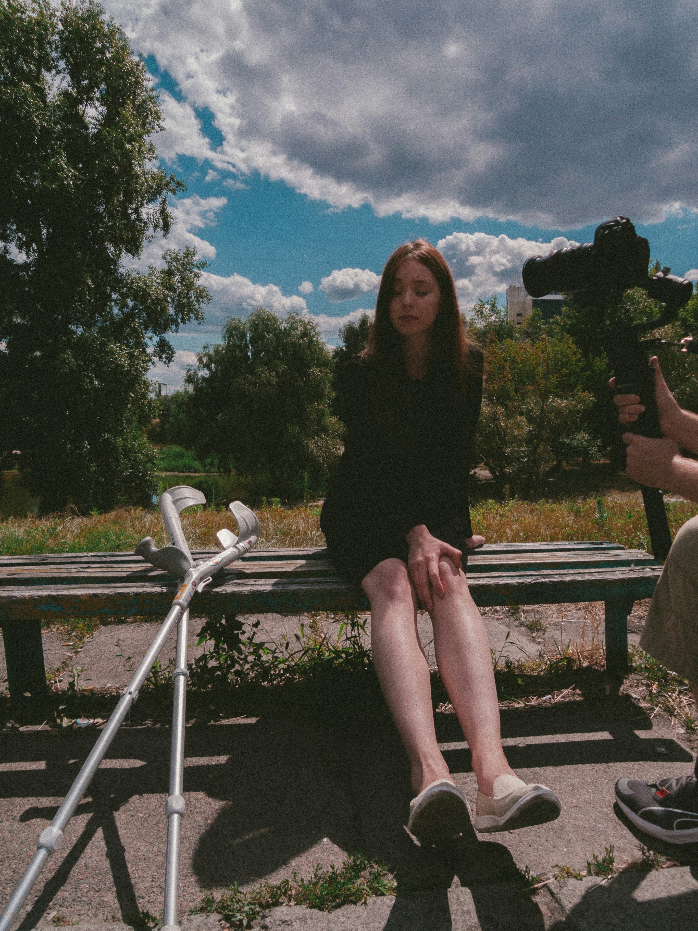A seated woman on a bench under a blue, clouded sky, with film gear nearby and a camera operator in frame; the scene centers on the subject with a cinematic, outdoorsy setup.