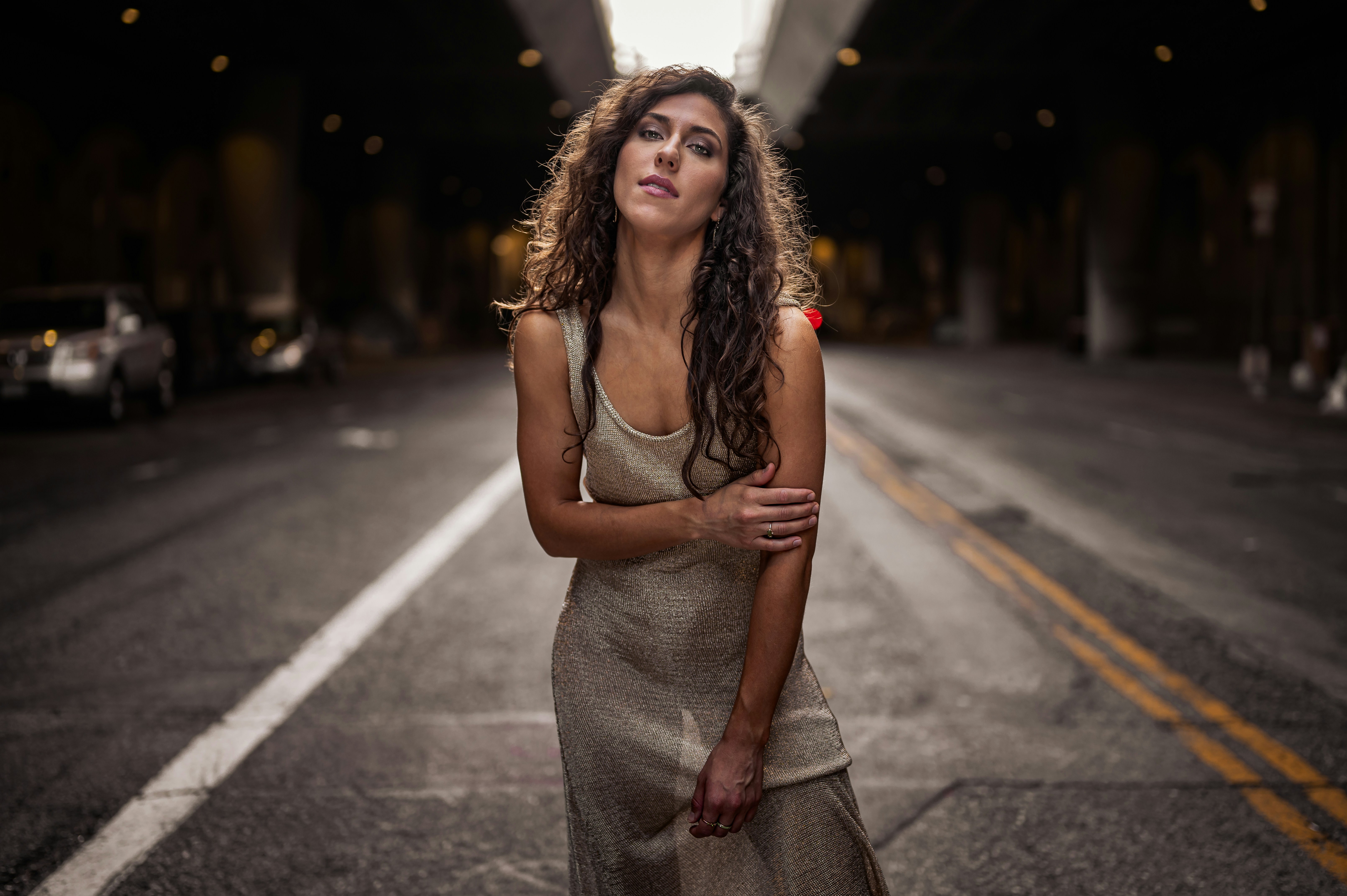 This striking portrait captures a woman in a shimmering gold dress standing confidently in an urban street setting. The composition uses leading lines of the road to draw attention to the subject, while the soft, ambient lighting creates a dramatic contrast with the darker background. The image's rich colors and dynamic atmosphere highlight both the elegance of the subject and the gritty charm of the cityscape.