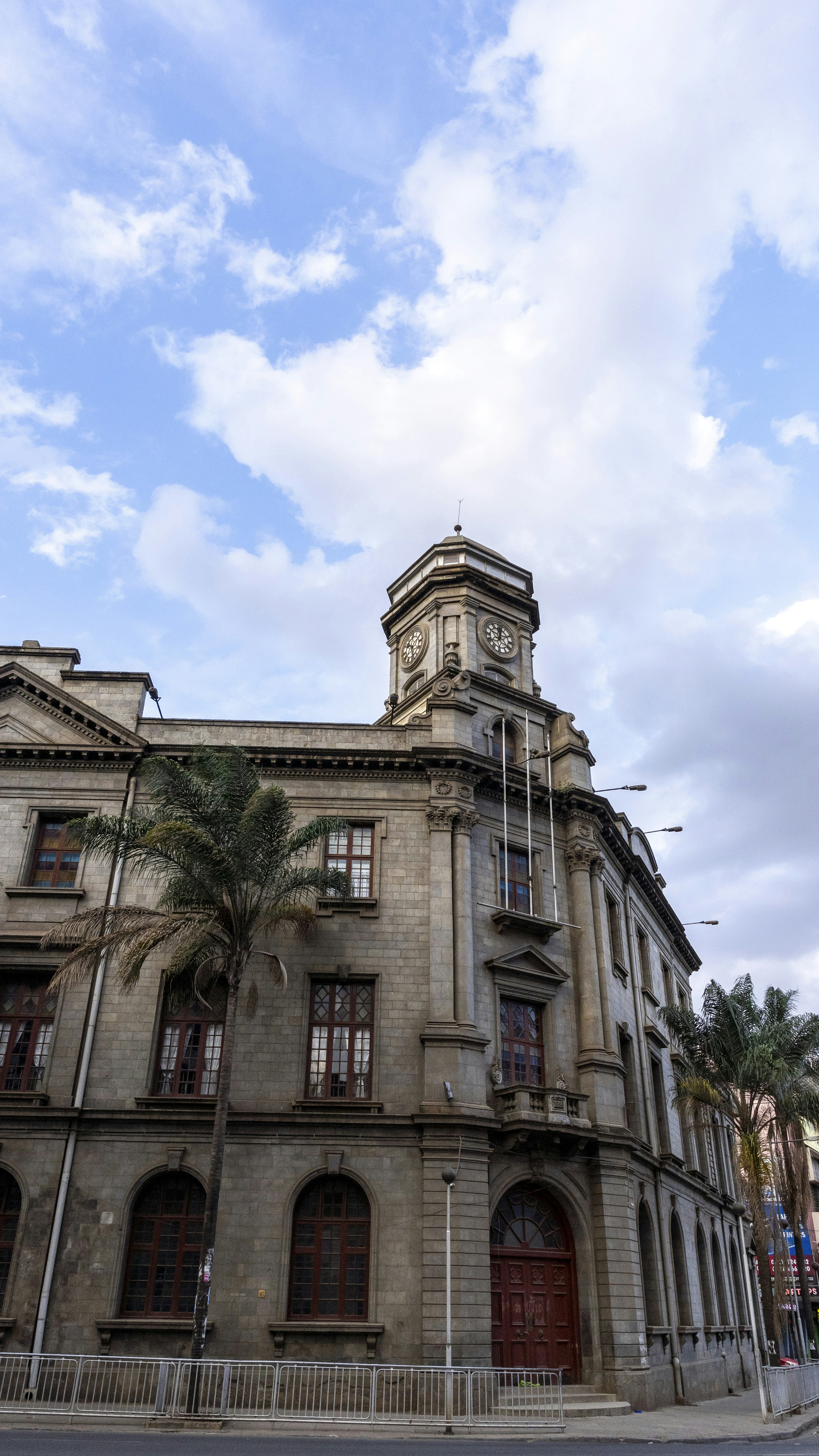 Historic building featuring intricate stonework and a prominent clock tower, framed by palm trees and a cloudy sky.