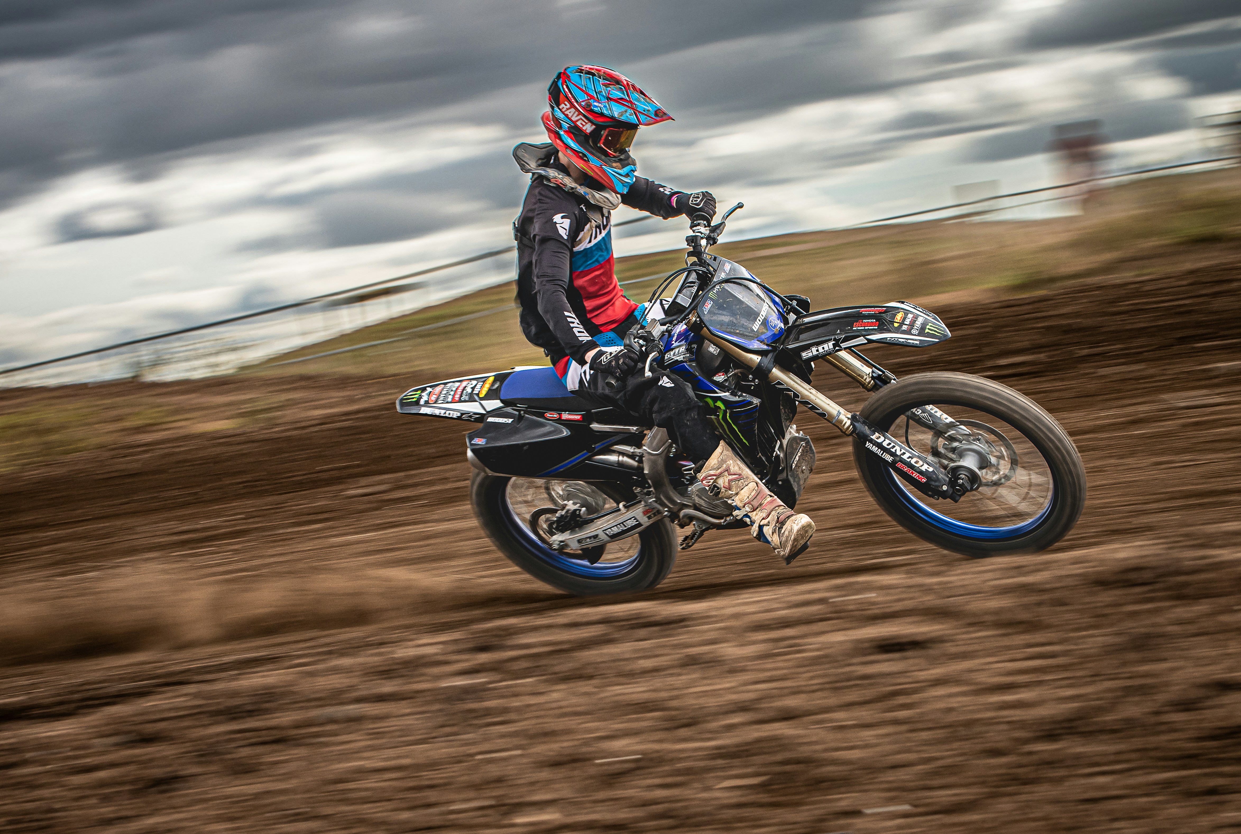 Motorcyclist in full gear racing on a dirt track with a blurred background and dramatic sky.