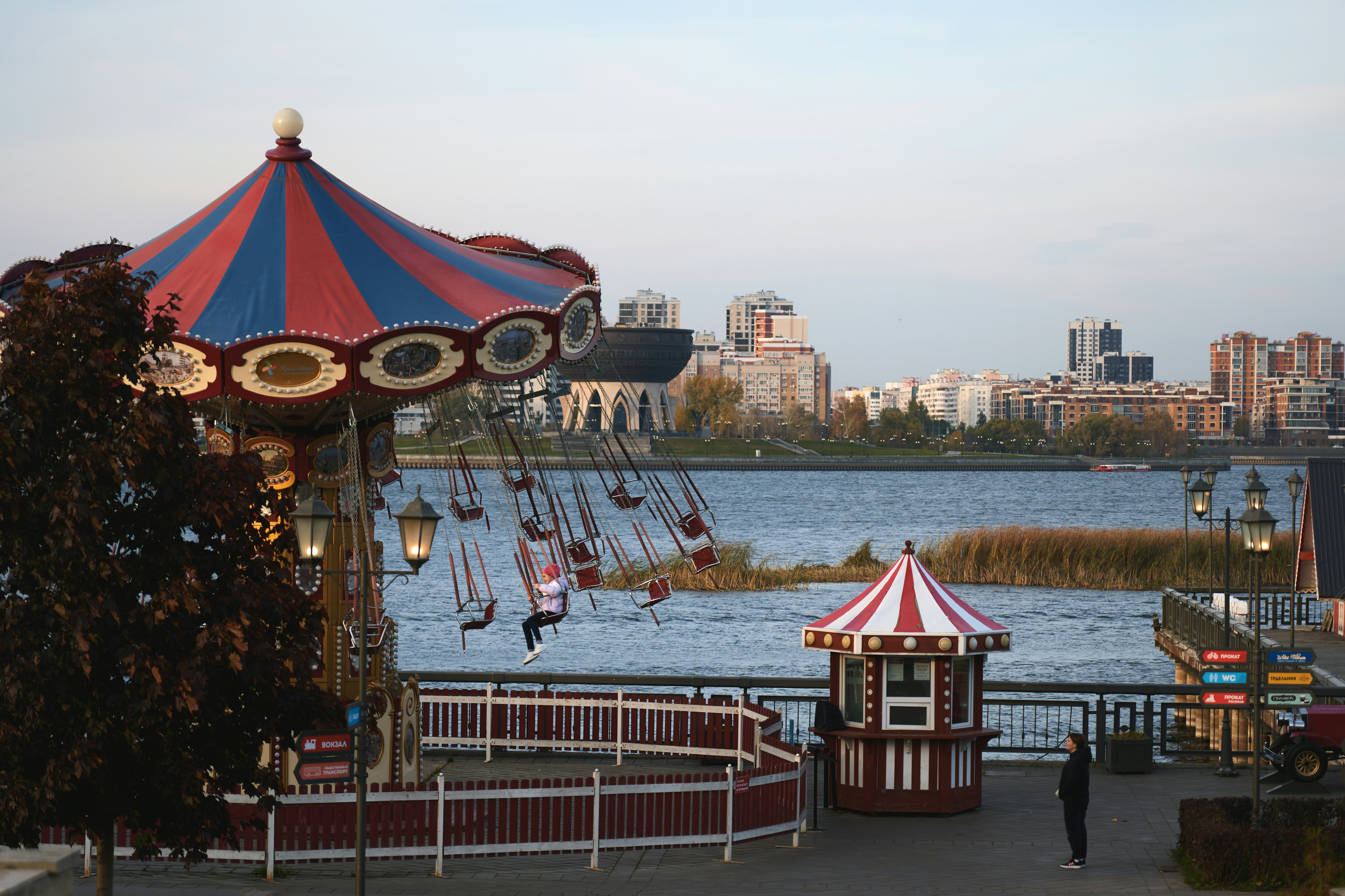 A vibrant carousel with colorful swings set against a serene lakeside backdrop, showcasing urban architecture in the distance.