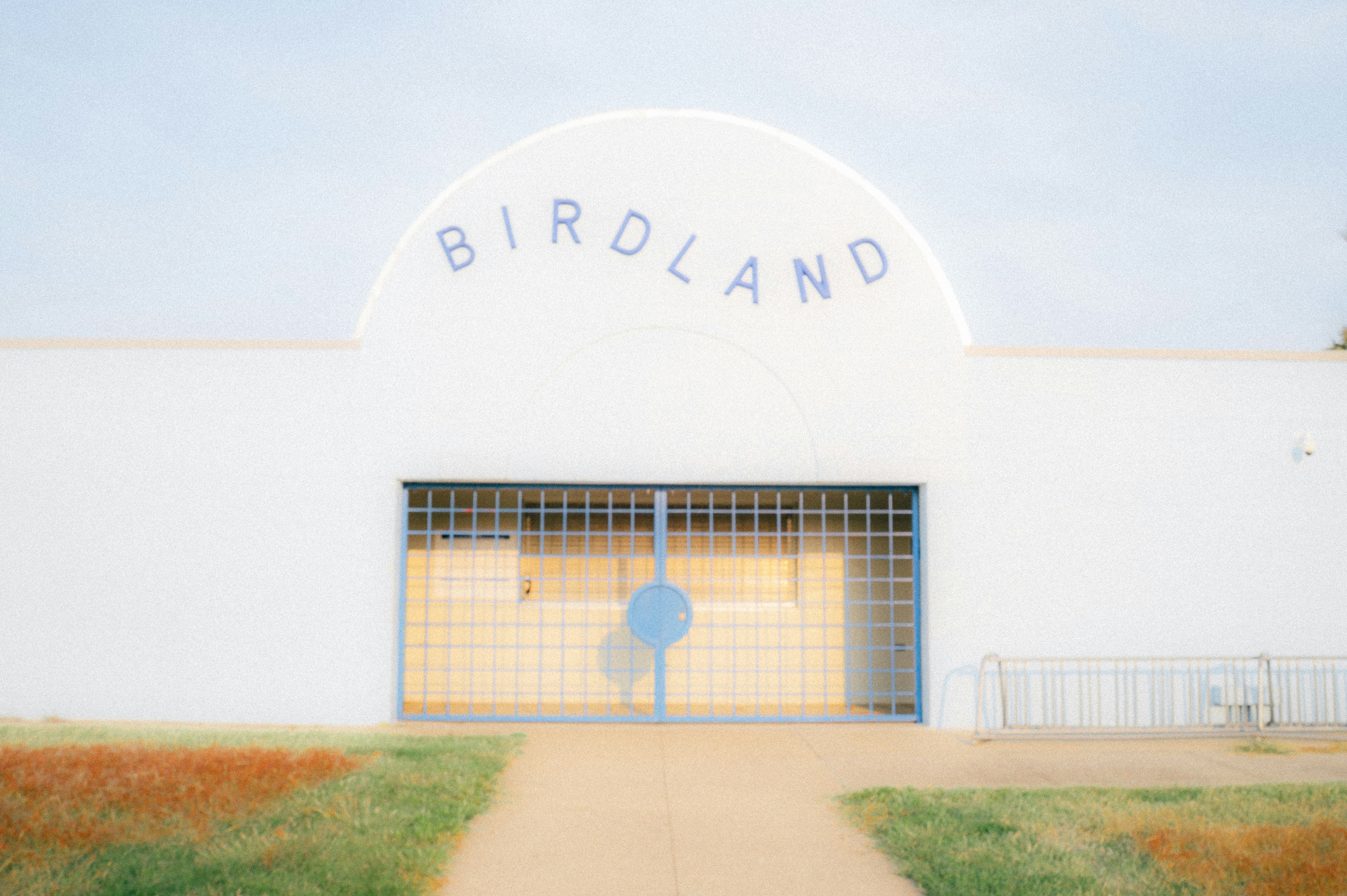 A minimalist facade of Birdland, featuring a prominent blue sign and a gated entrance. The soft color palette enhances the serene atmosphere.