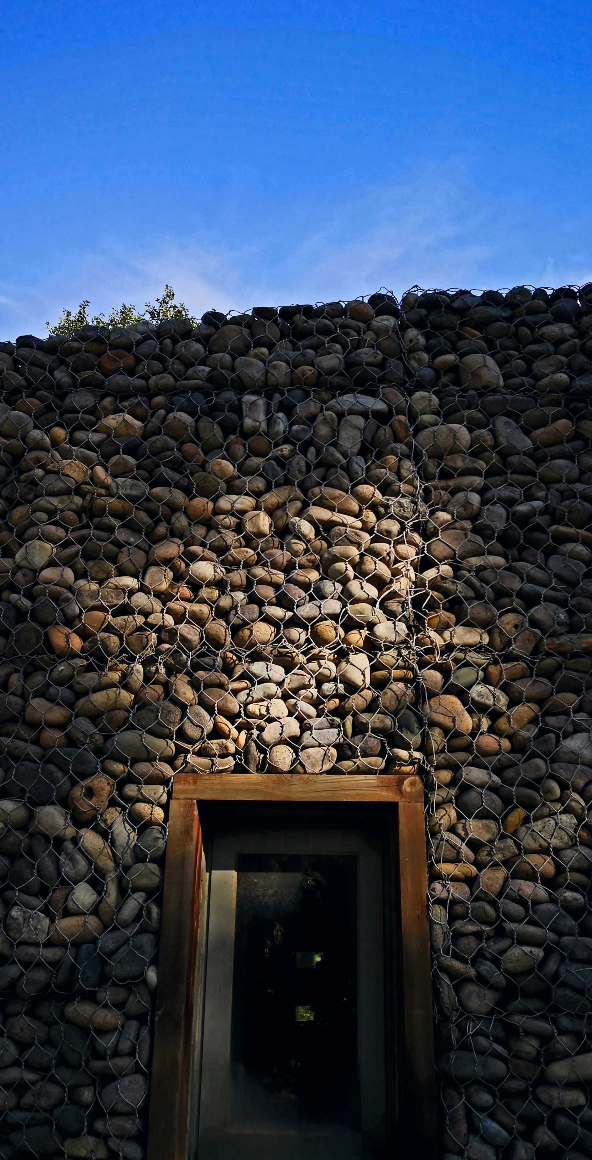 Textured stone wall with a wooden door framed at the center, under a clear blue sky. The arrangement of stones creates a natural aesthetic.