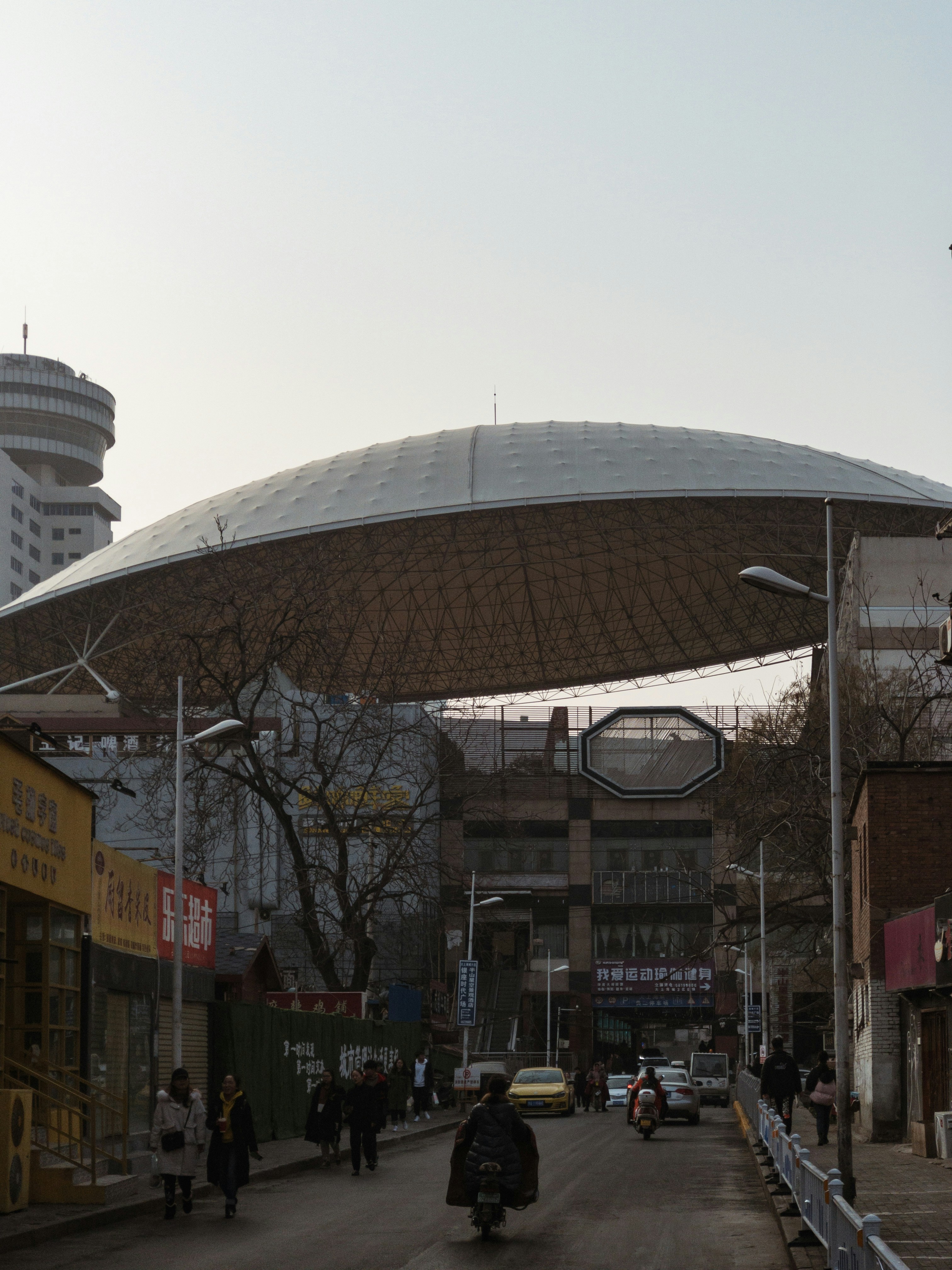 A large, modern architectural canopy covers a bustling urban street, with buildings and vehicles visible beneath it.