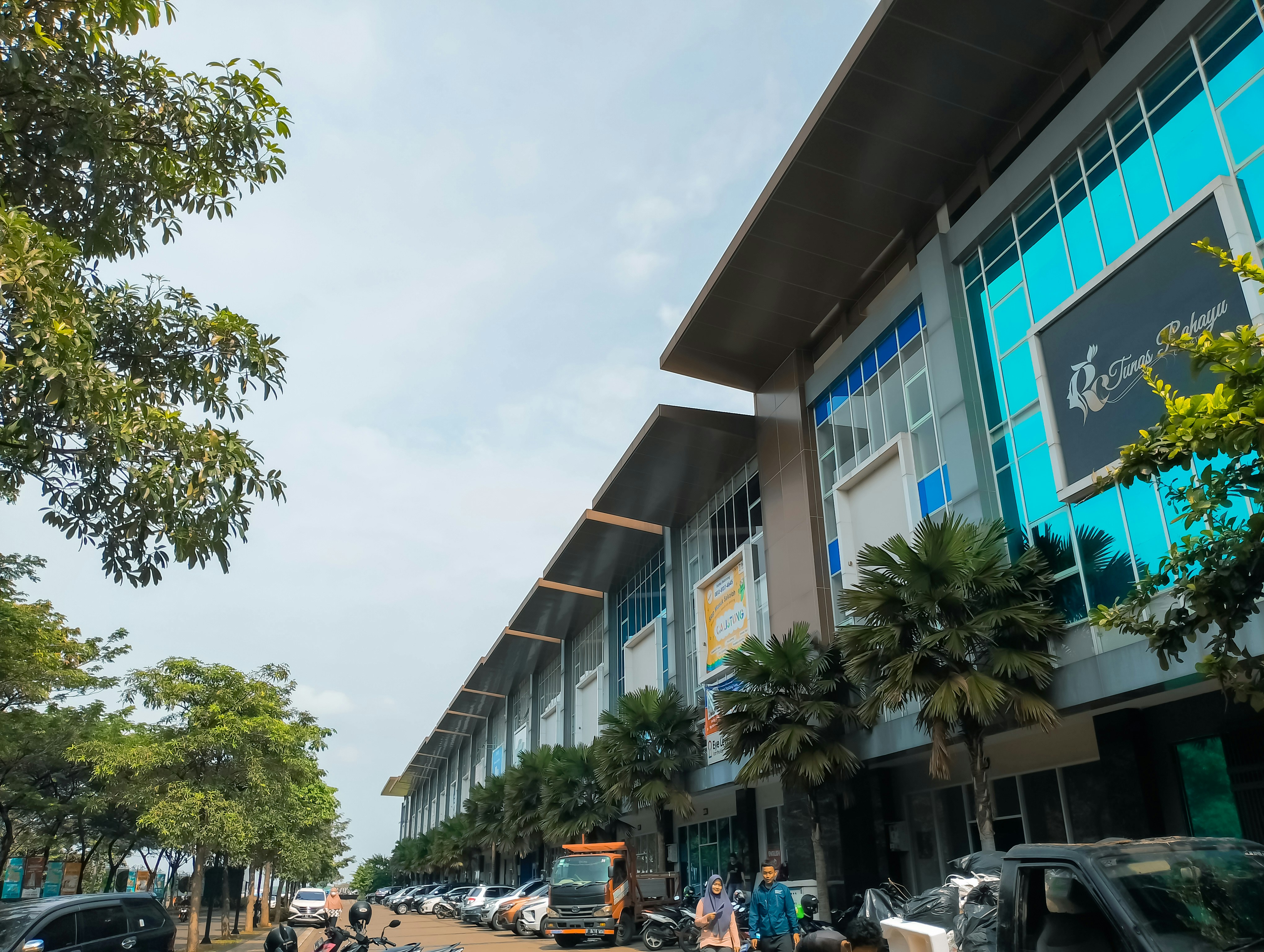Contemporary building with glass facade and palm trees lining a busy street under a partly cloudy sky.