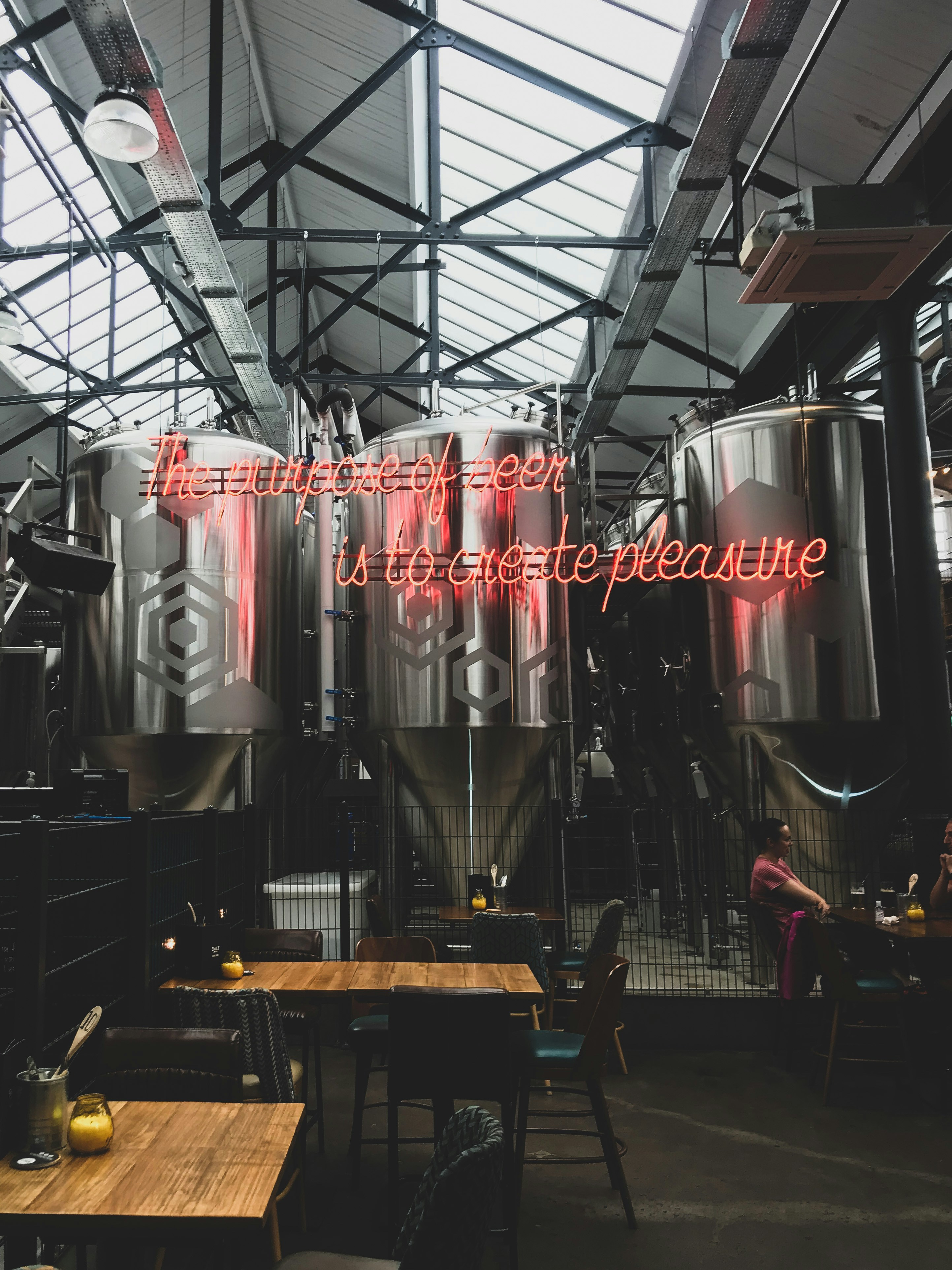 Illuminated neon sign reading 'The purpose of beer is to create pleasure' in a modern brewery setting with metallic brewing tanks.