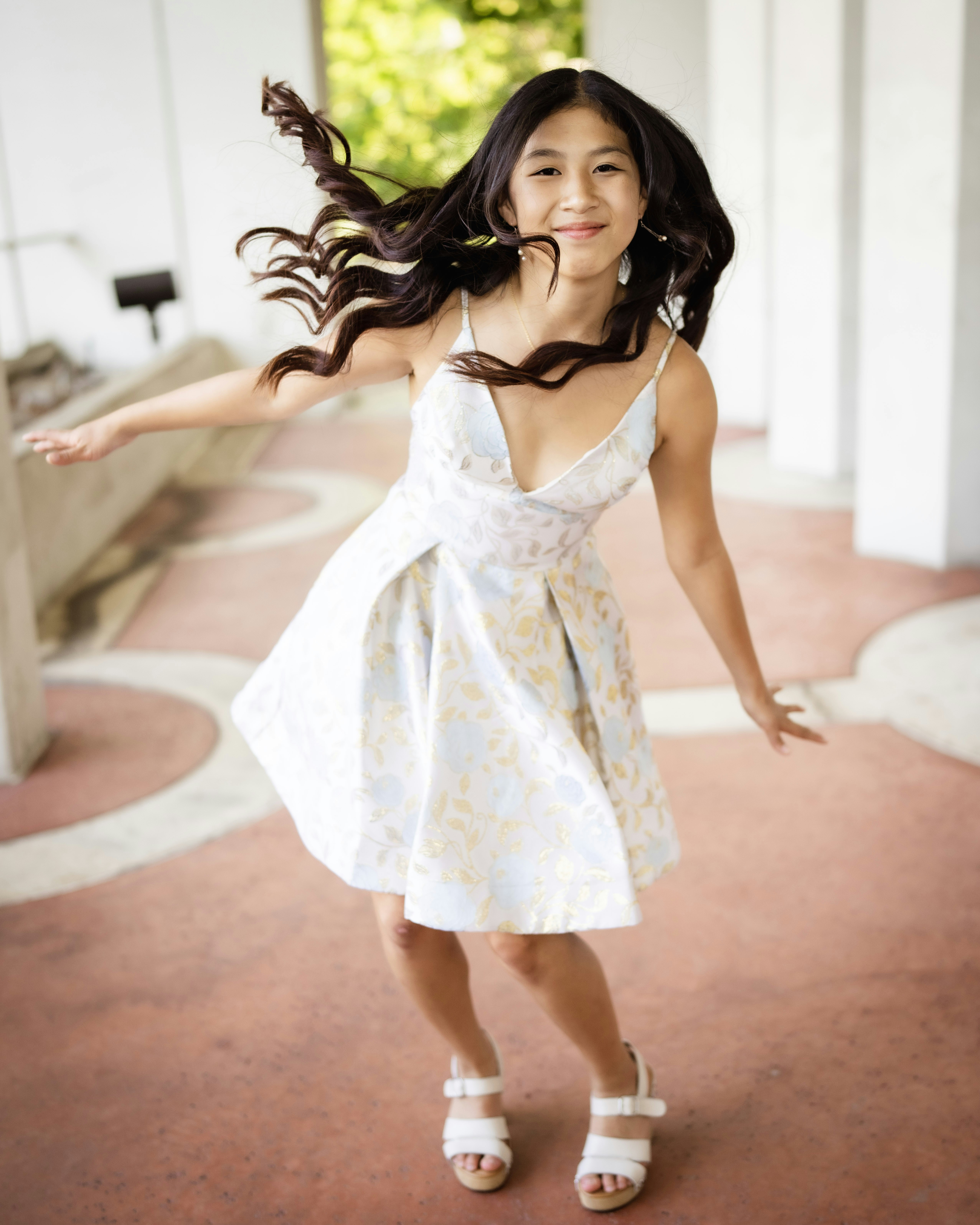 Teenage girl spinning in a floral dress on a sunlit patio.