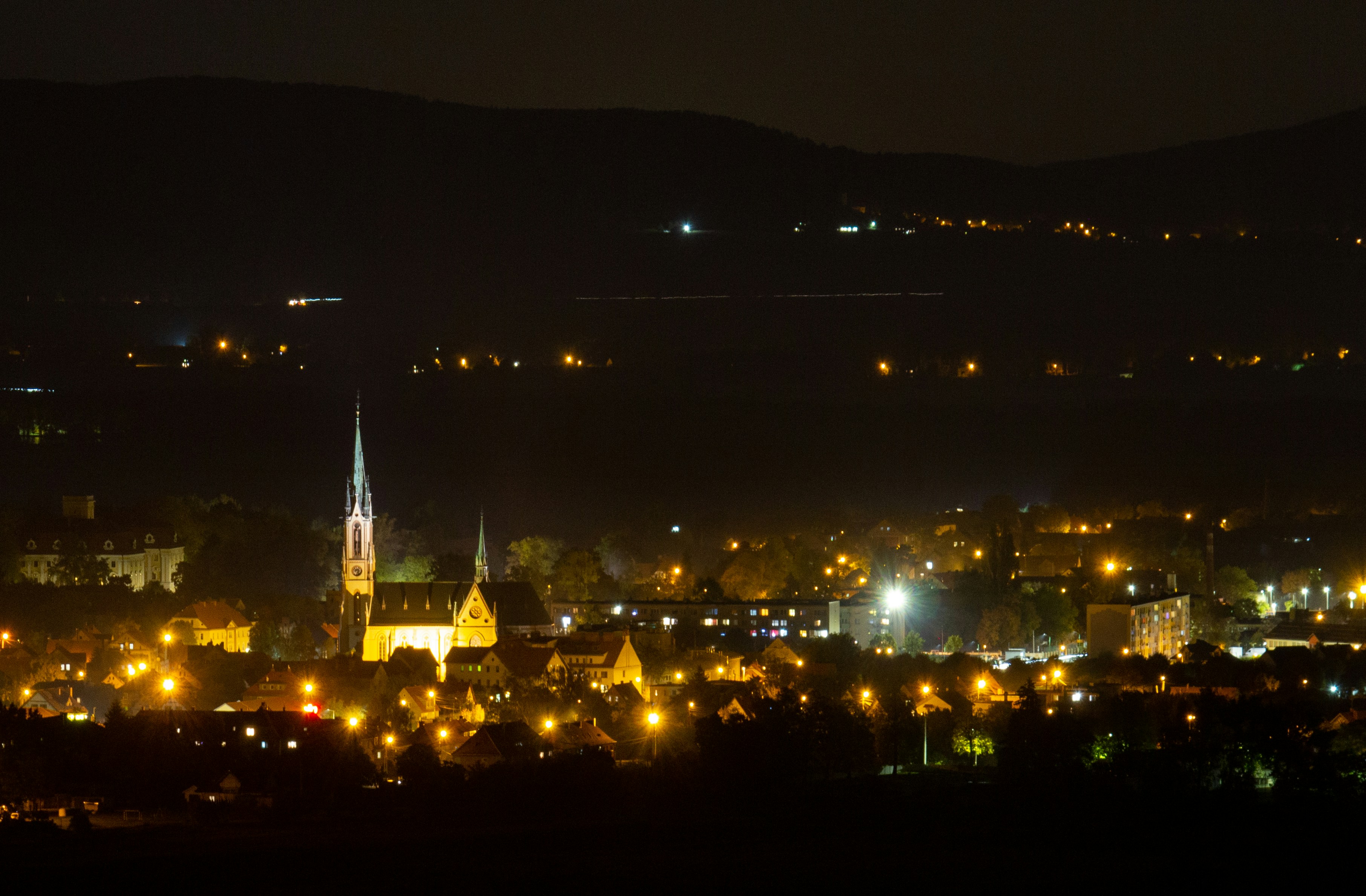 Nighttime cityscape with illuminated church spires against a backdrop of dark hills.