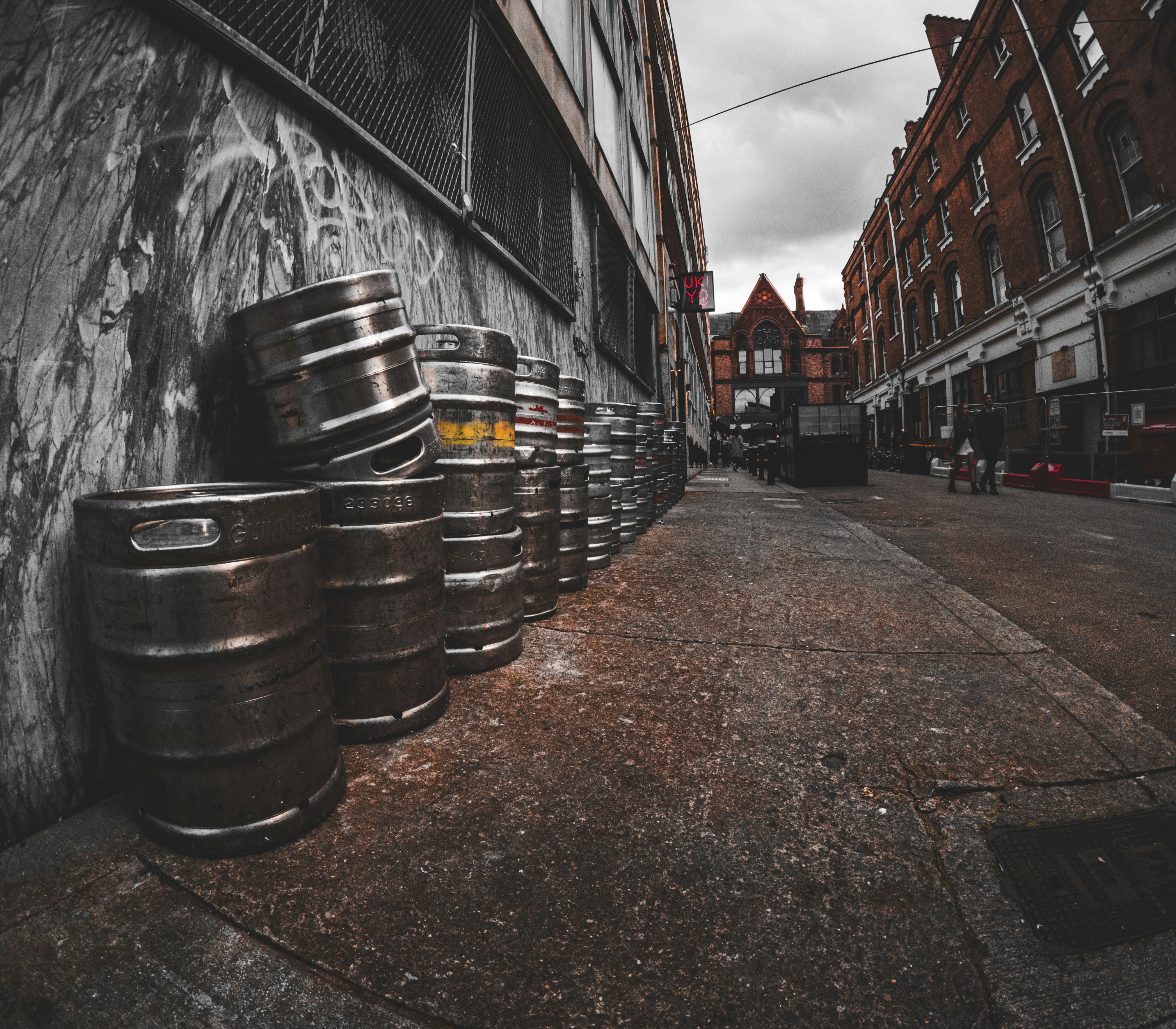 Rows of metal beer kegs line a gritty city sidewalk, guiding the eye toward a distant urban street. This street photograph emphasizes texture and perspective in an industrial setting.