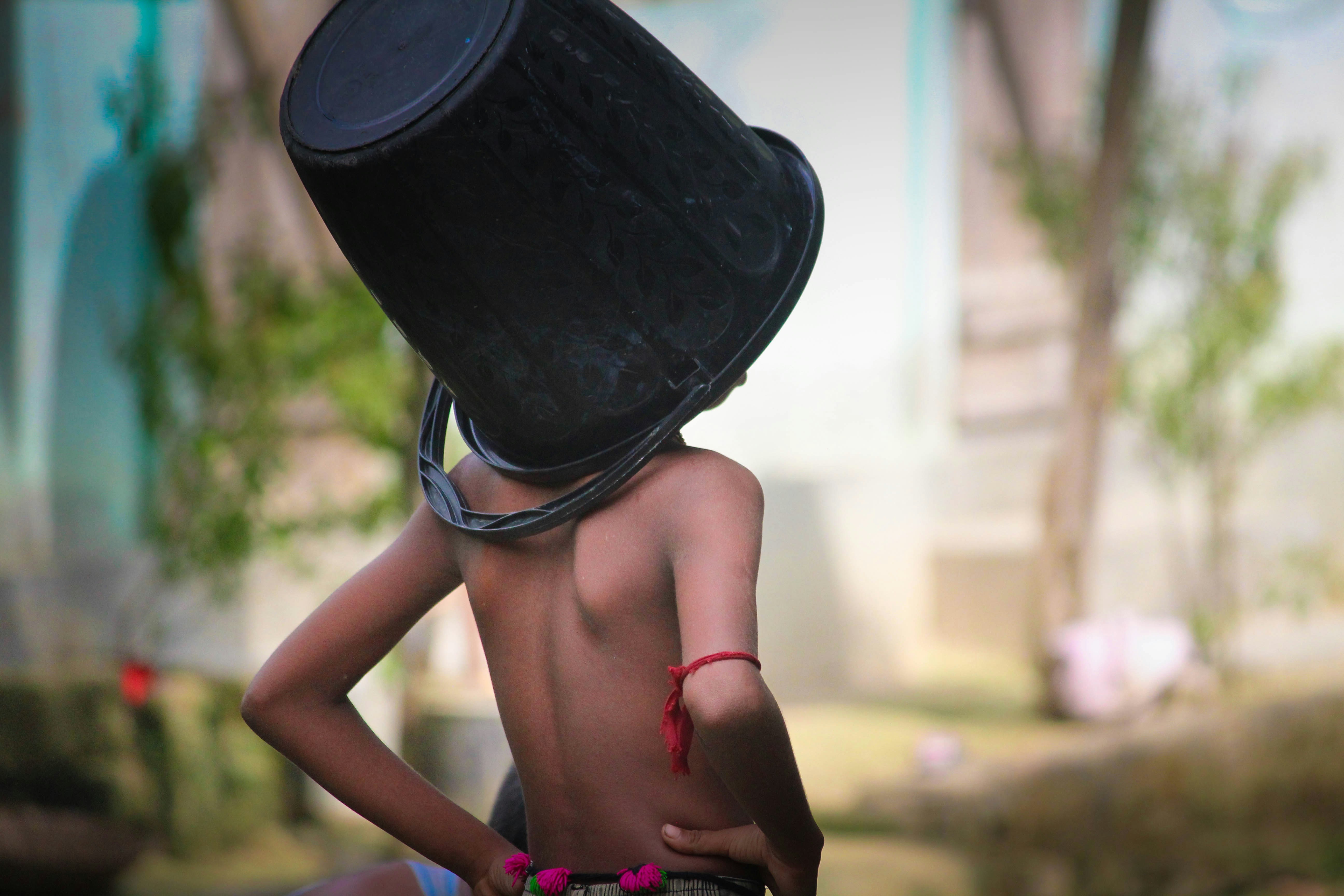 A shirtless child with a bucket on their head strikes a playful pose in an outdoor setting; the scene emphasizes whimsy and summer play, with soft background blur. Note: this is a candid stylized shot featuring a prop in the foreground.