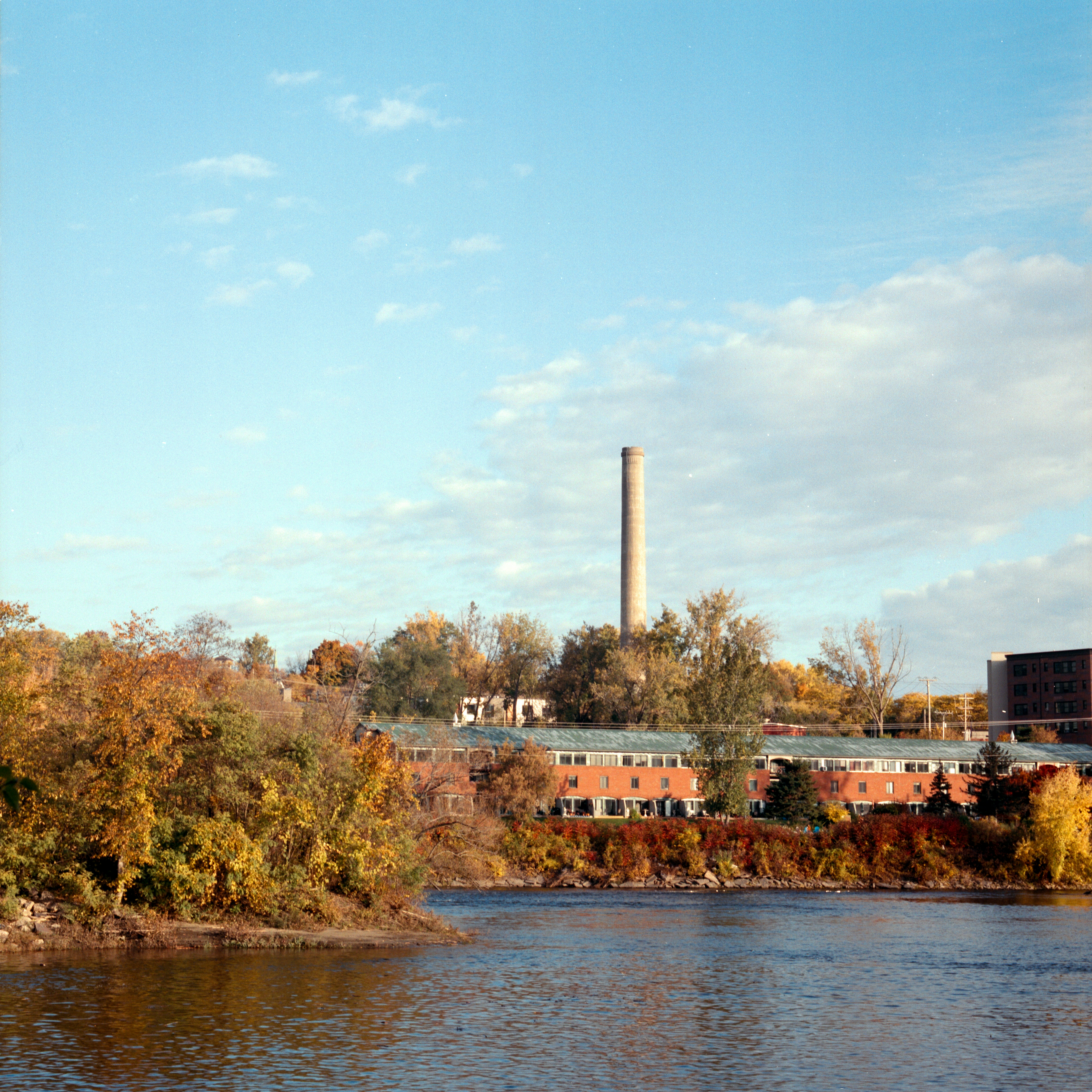 A serene riverside view showcasing vibrant autumn foliage alongside a historic industrial structure and a tall smokestack. The tranquil water reflects the colorful landscape.