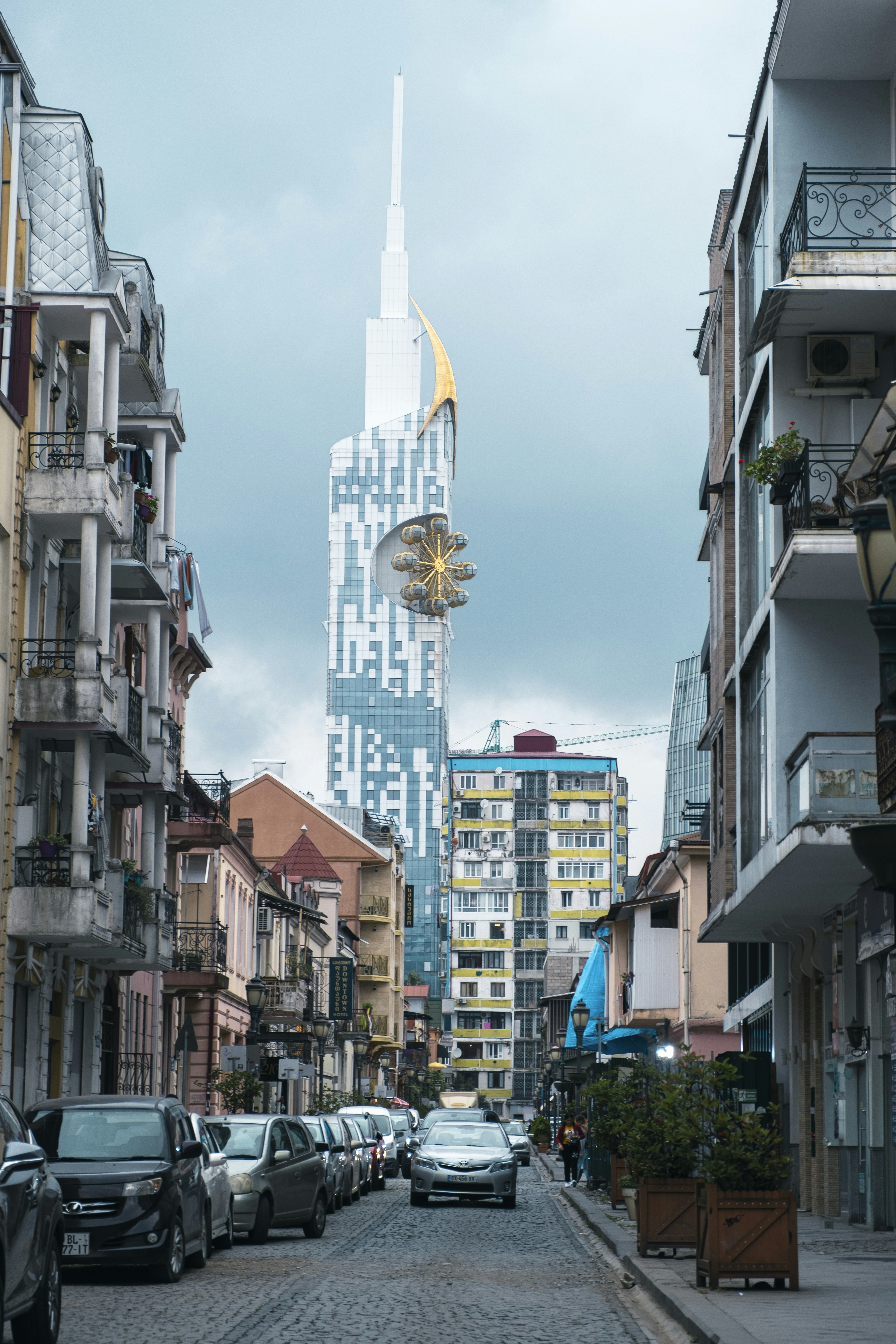 A bustling street lined with traditional buildings leads to a striking modern skyscraper, showcasing a blend of architectural styles. The scene captures the essence of urban life in a vibrant city.