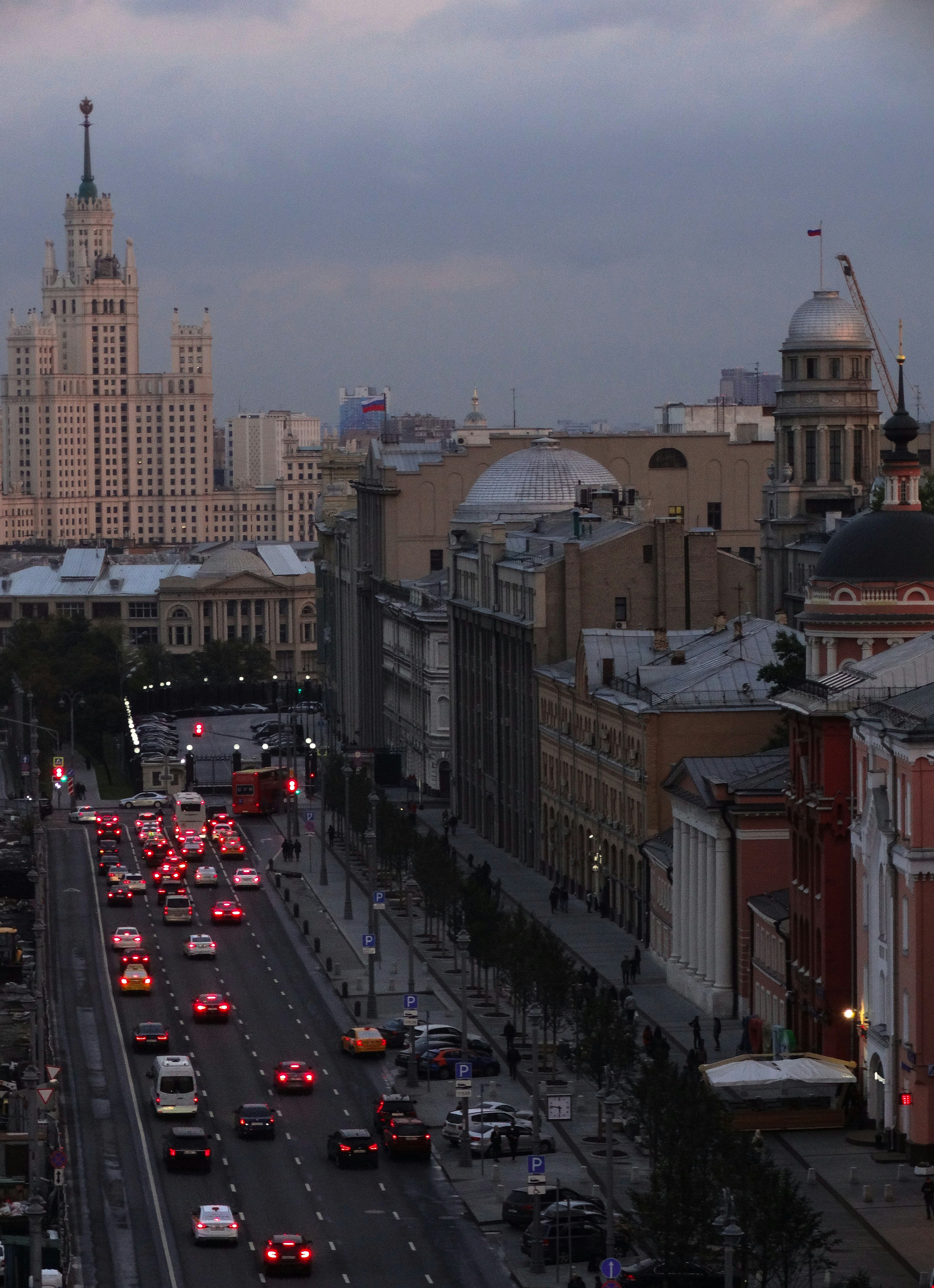 Cityscape photograph of a busy urban avenue lined with historic buildings at dusk, with a prominent left-spired tower anchoring the skyline.