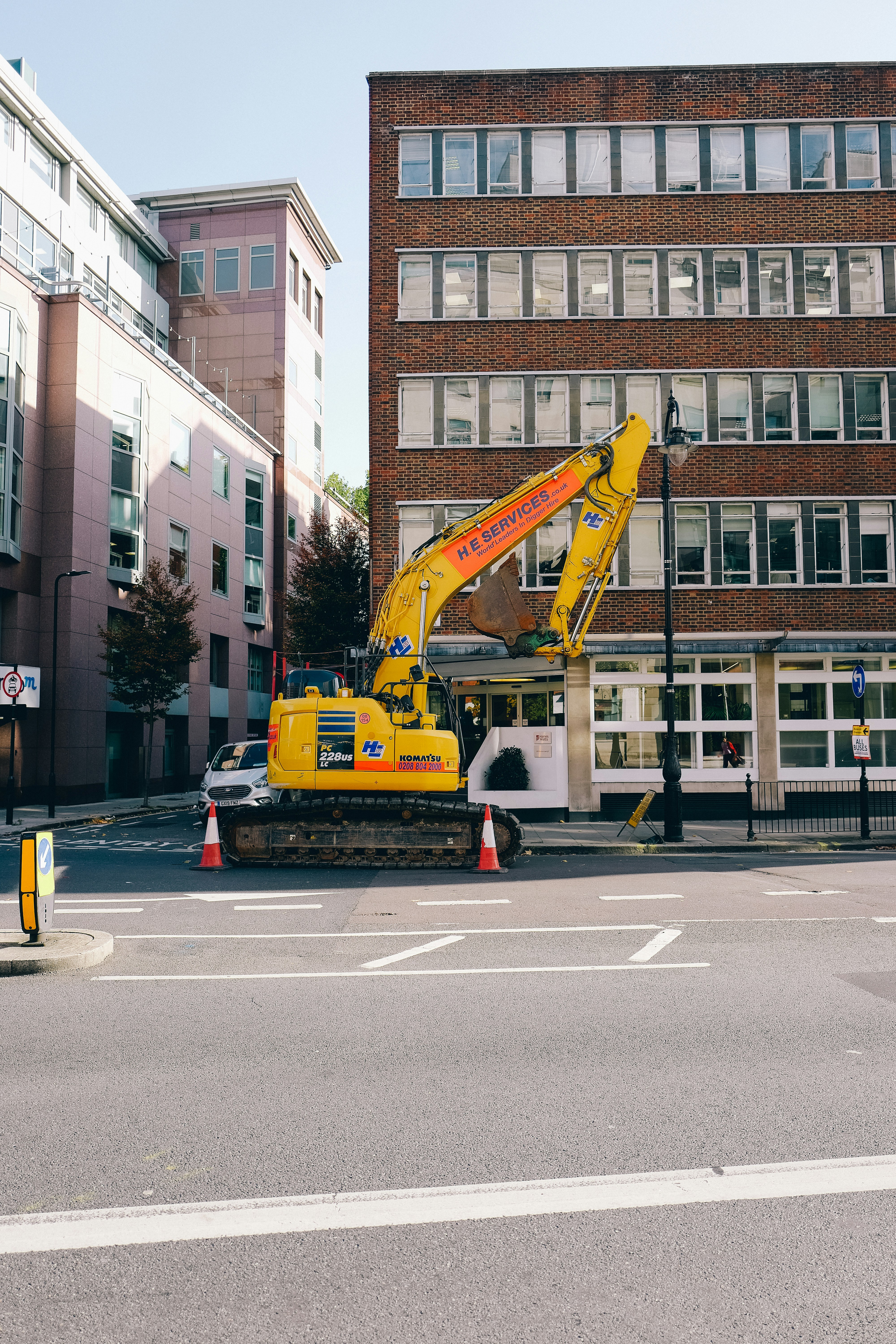 Excavator parked on a city street, poised for construction work near a brick building. Traffic cones and urban structures frame the scene.