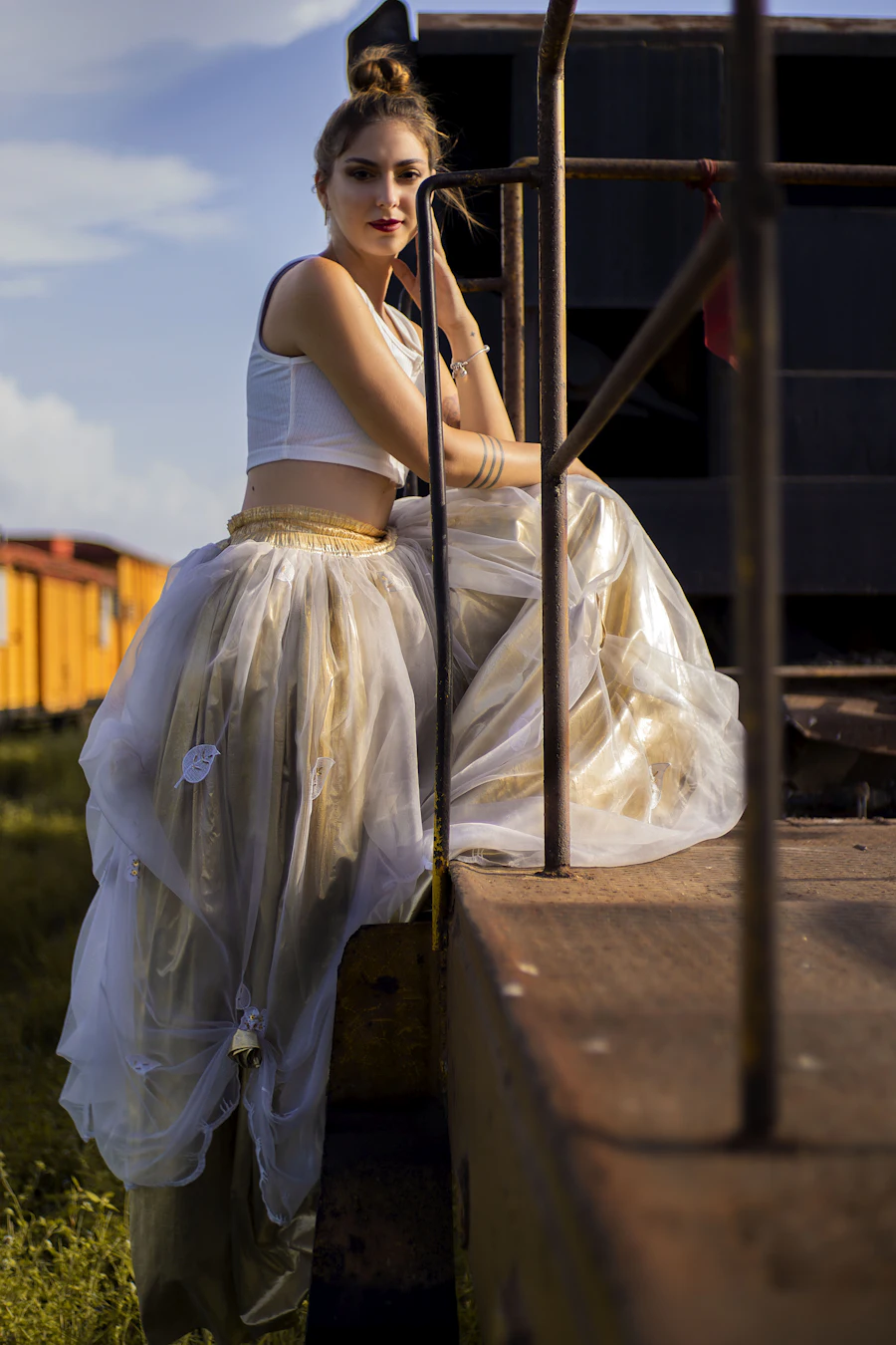 Young woman in a white crop top and sheer flowing skirt sits on rusty industrial stairs outdoors, framed by metal railings against a blue sky.