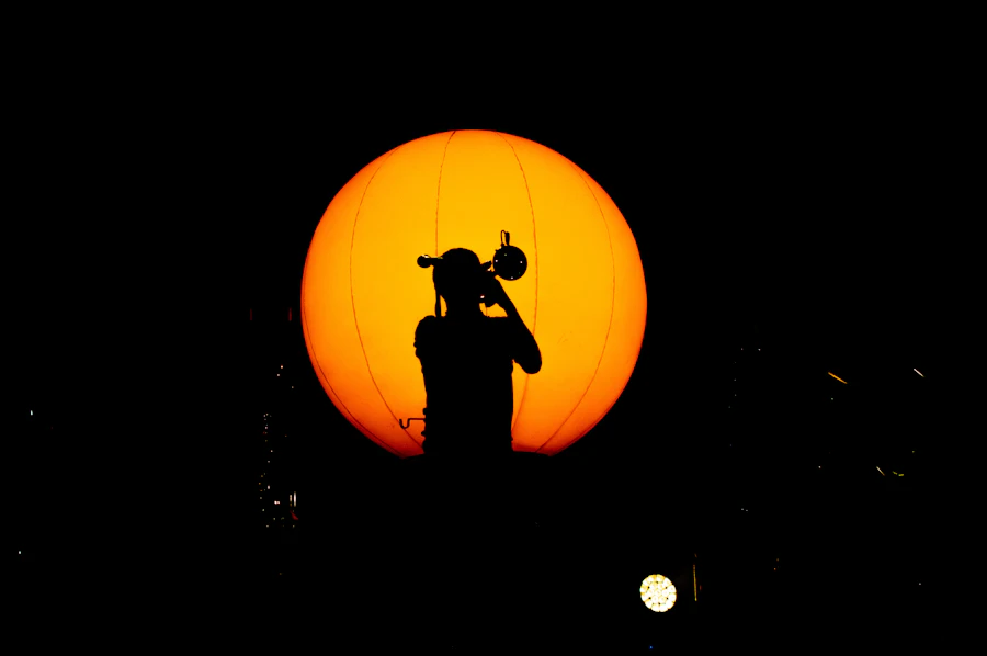 A silhouetted performer stands in front of a glowing orange circular light, with a small spotlight below. The scene is nearly black except for the bright orb and the figure's outline.