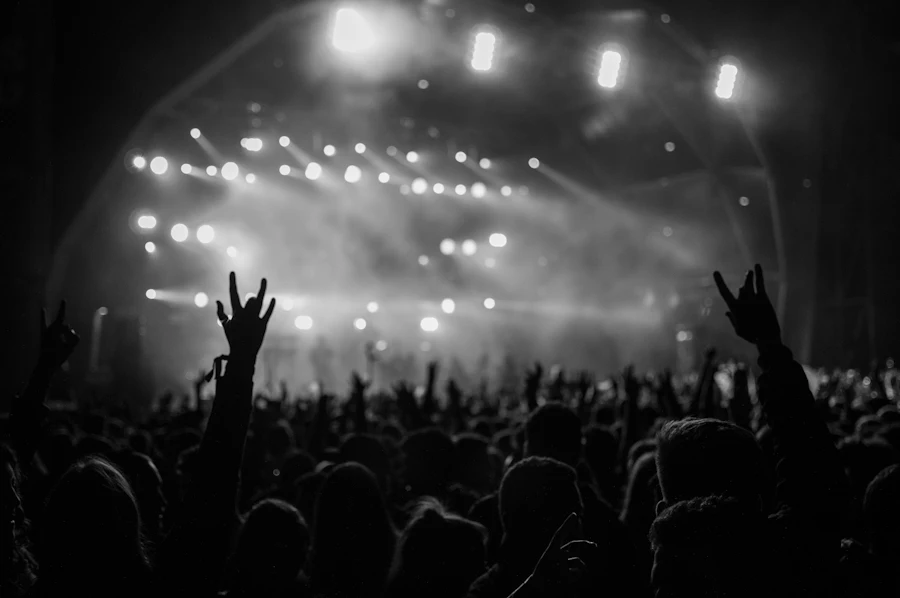 A packed crowd cheers with raised hands in front of bright stage lights and haze at a live concert.