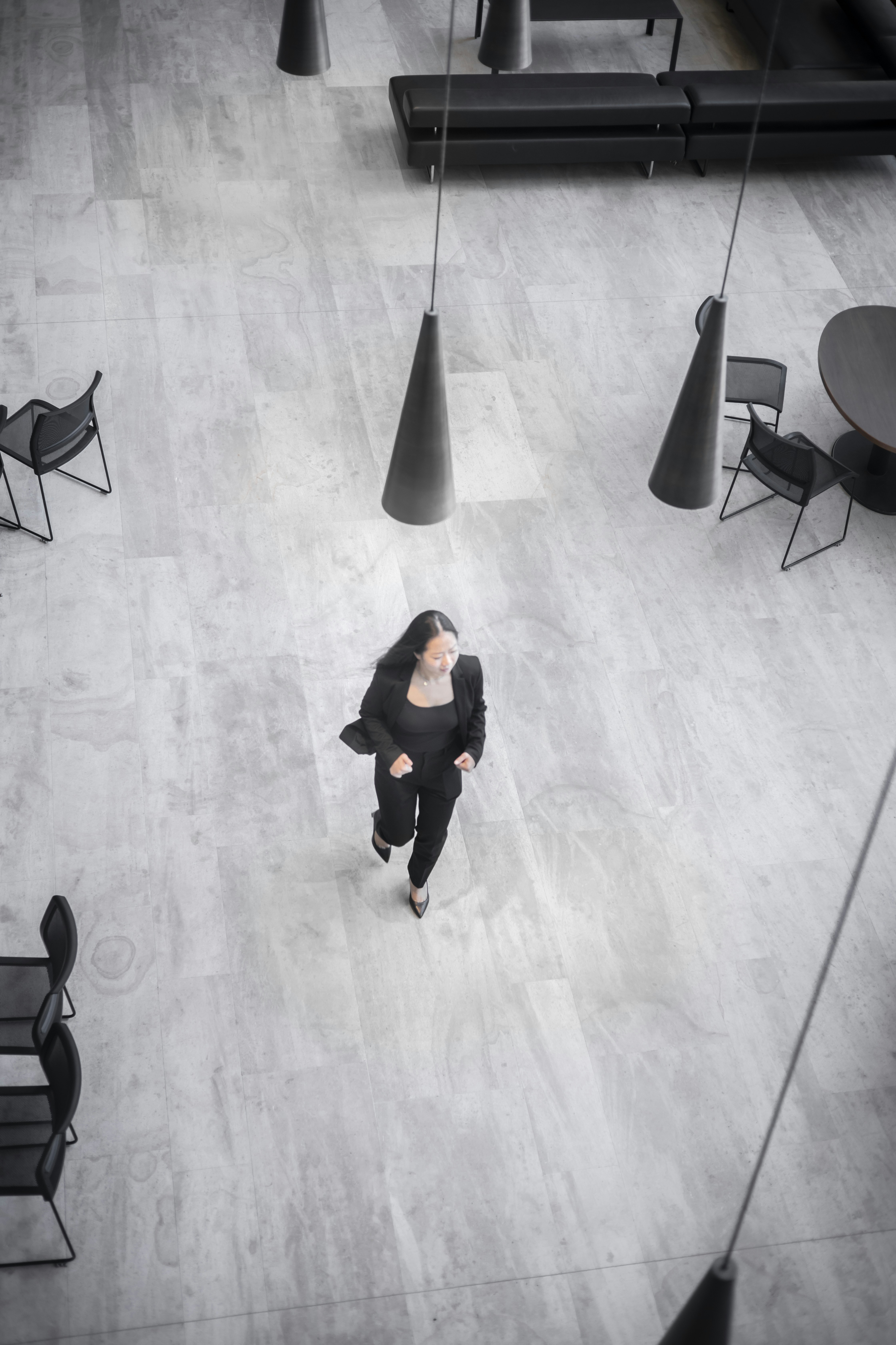 Overhead view of a woman in black attire walking briskly across a spacious, light-toned marble floor.