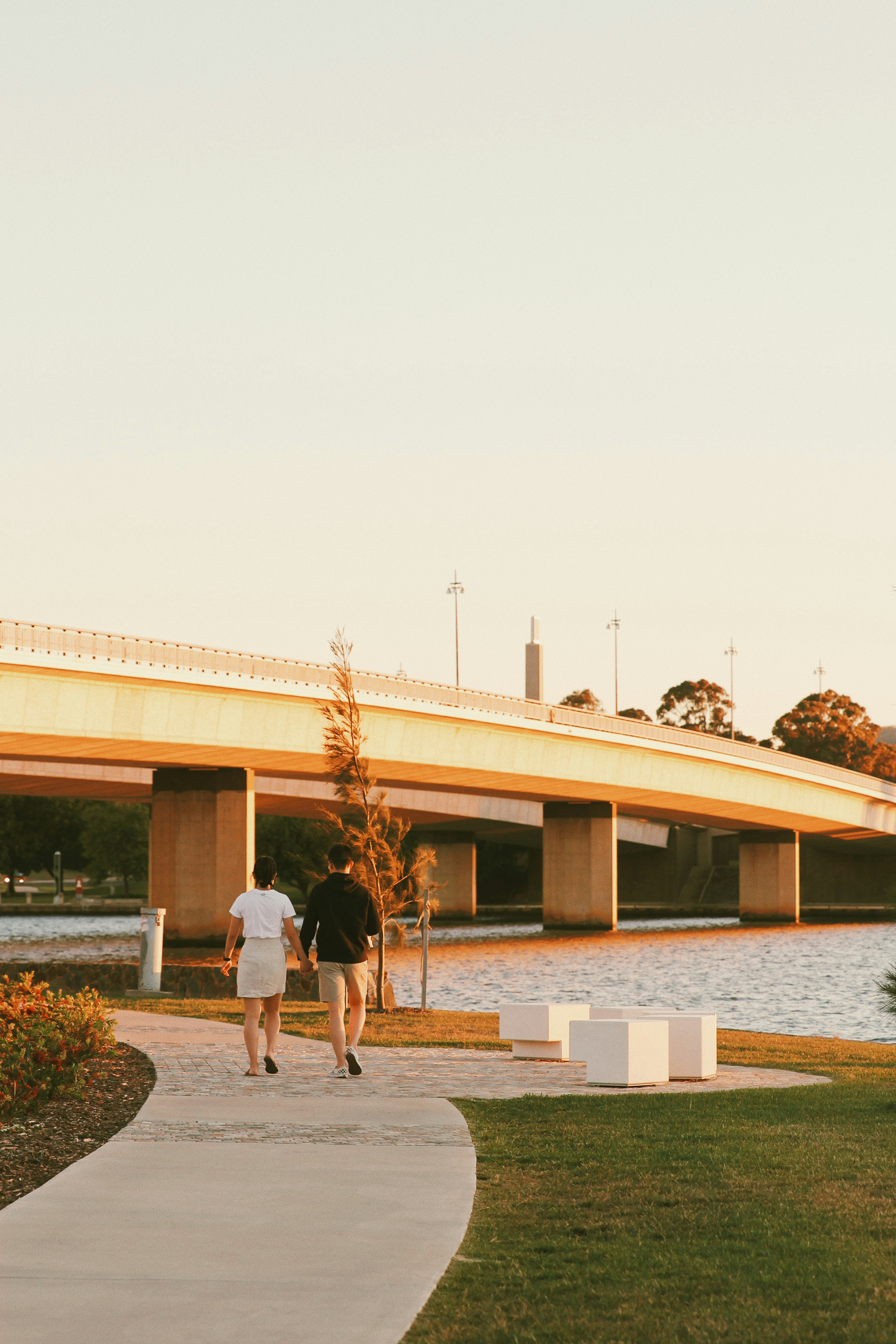 Couple walking hand-in-hand along a riverside path, with a bridge arching gracefully above them during golden hour.