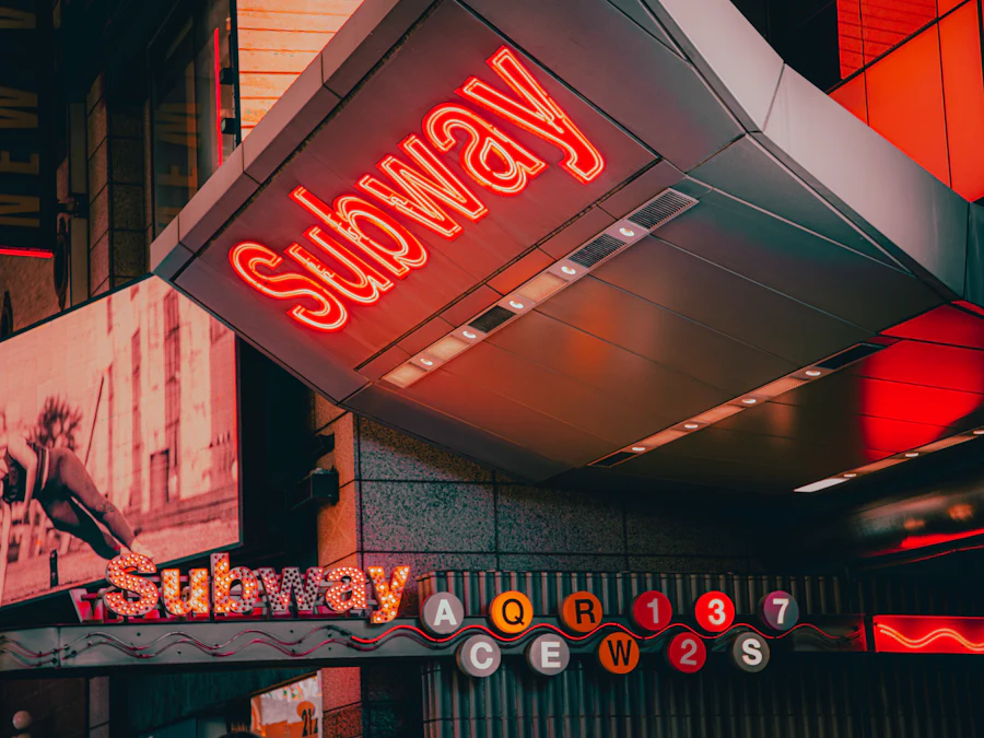 A glowing red subway sign hangs over a New York City entrance, with route circles and a billboard visible below. The scene is lit by vivid neon and street reflections.