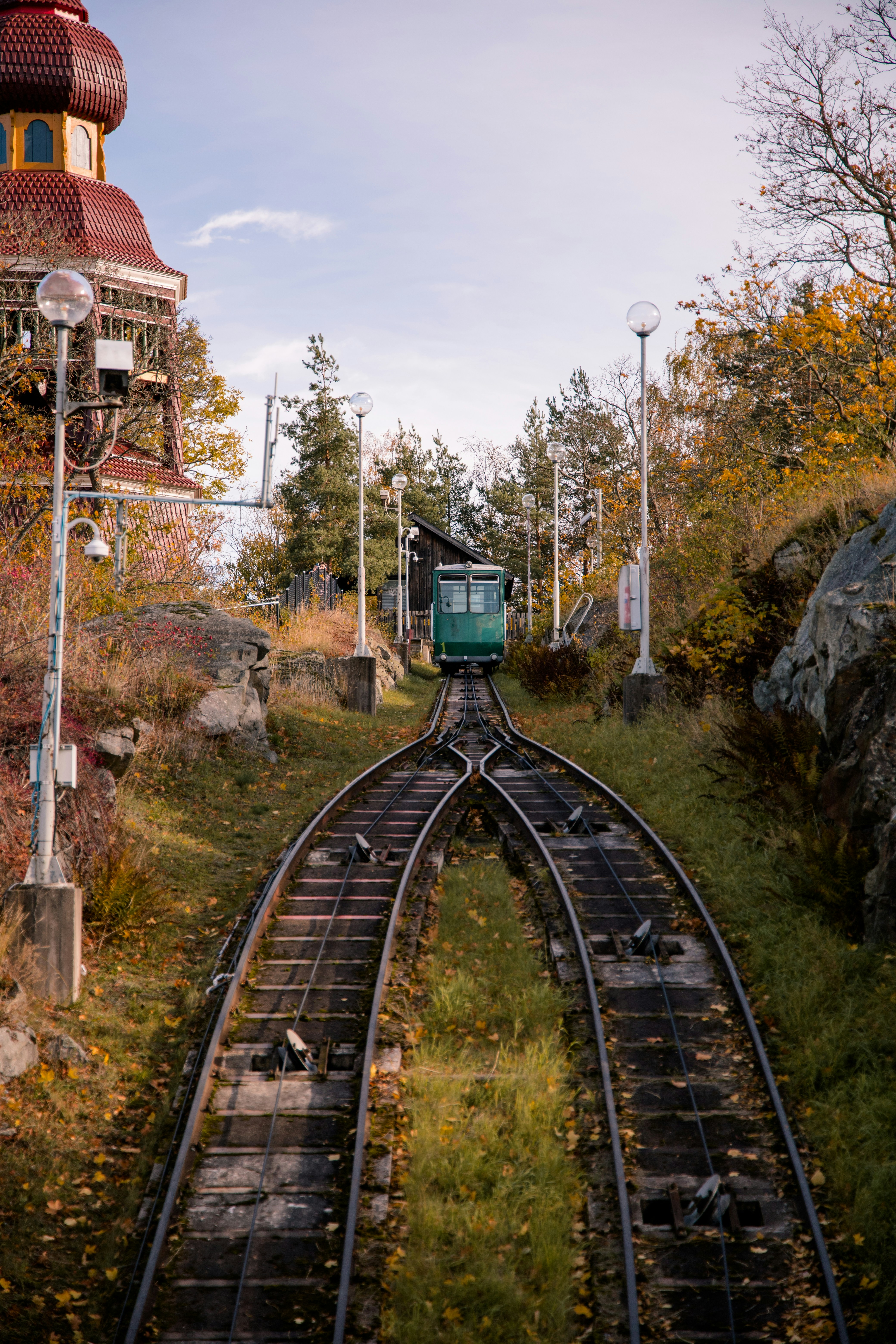 A narrow-gauge railway curves toward a small green carriage against a late-autumn landscape with trees and a red-roofed building on the left. The scene captures a quiet moment at a compact station, highlighting the tracks and surrounding foliage.