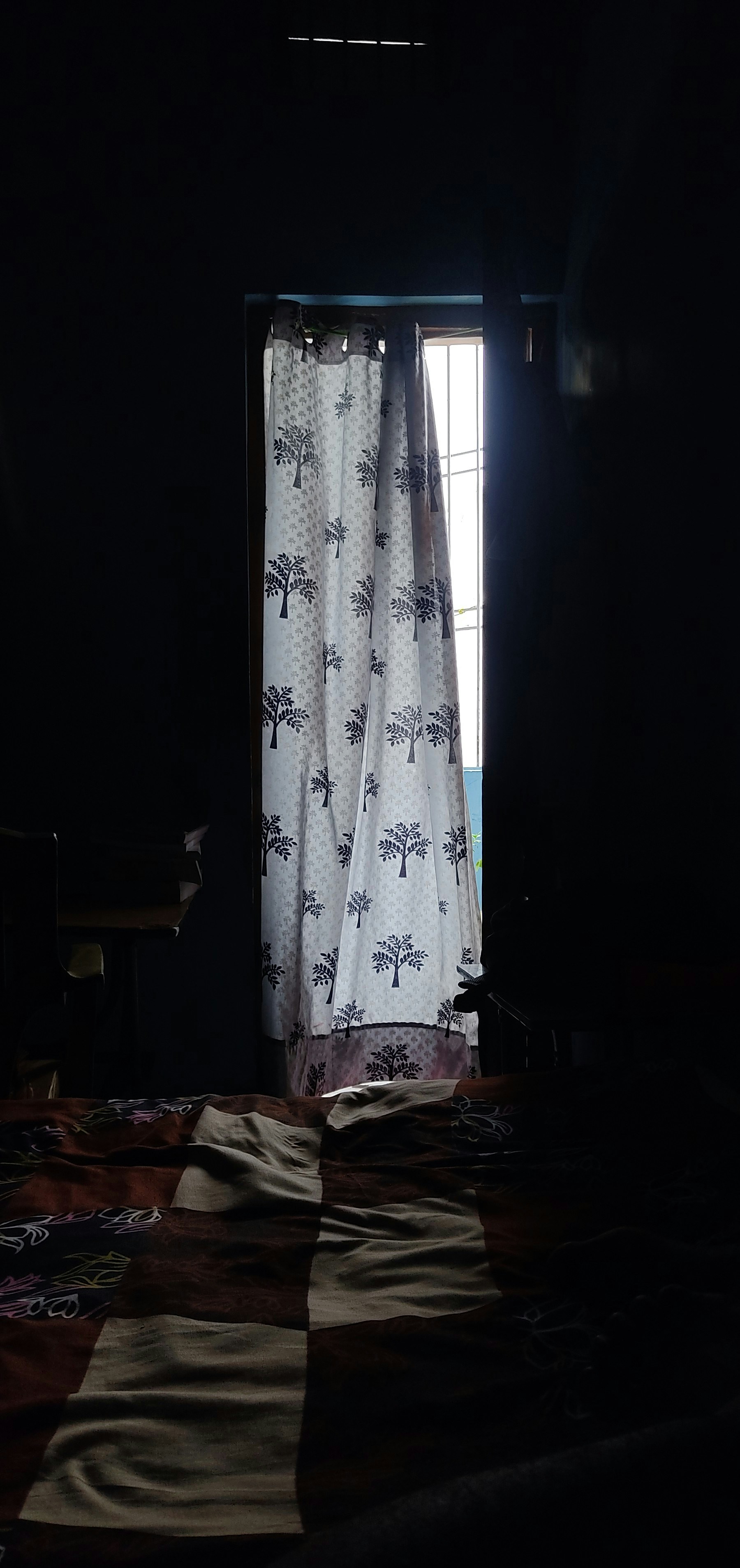 Sheer curtain adorned with tree motifs sways gently in front of an open window, allowing soft light to filter into a dimly lit room.