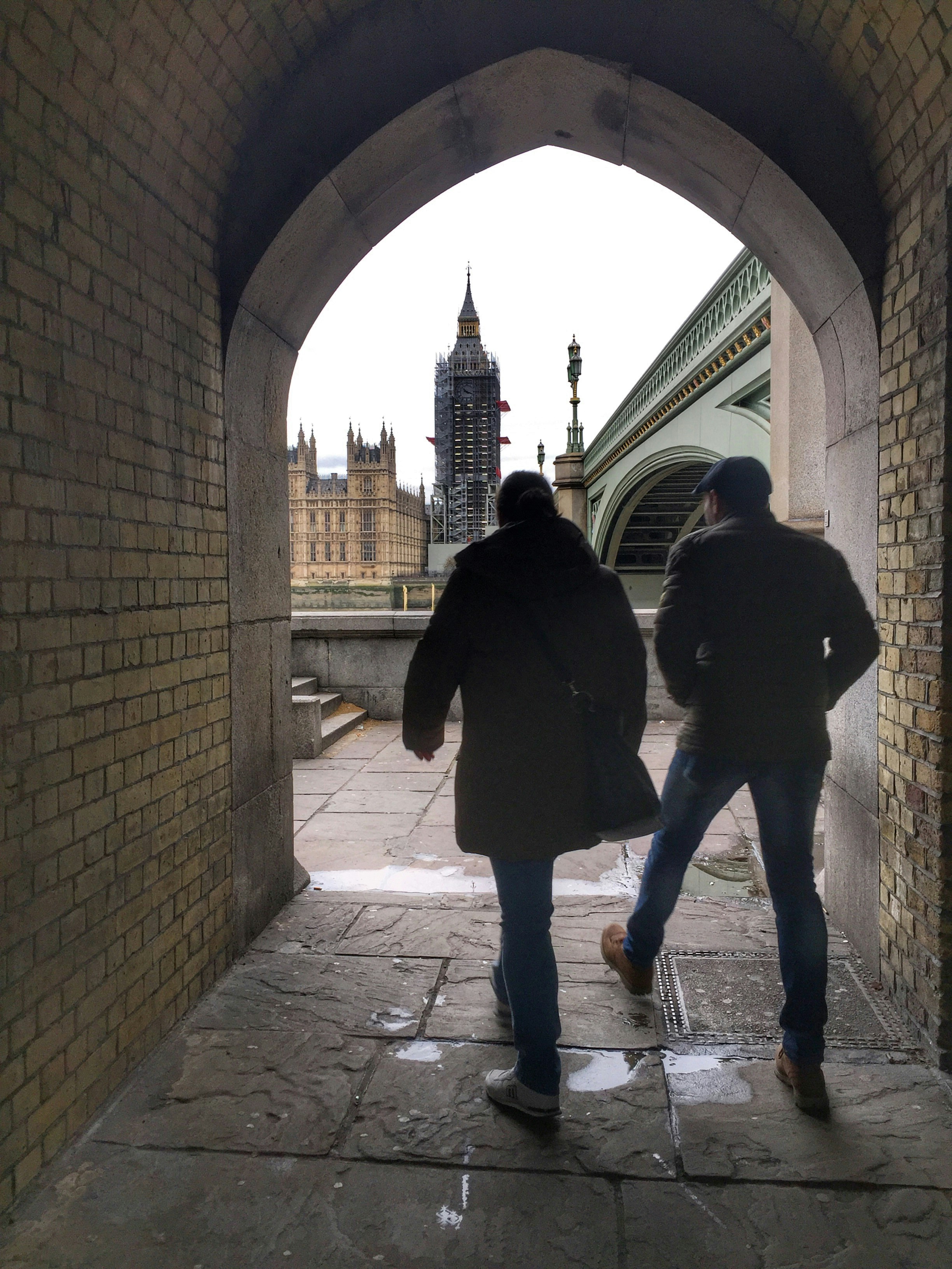 Two figures walking through an archway towards the iconic Big Ben and Westminster Bridge, framed by historic brickwork. 