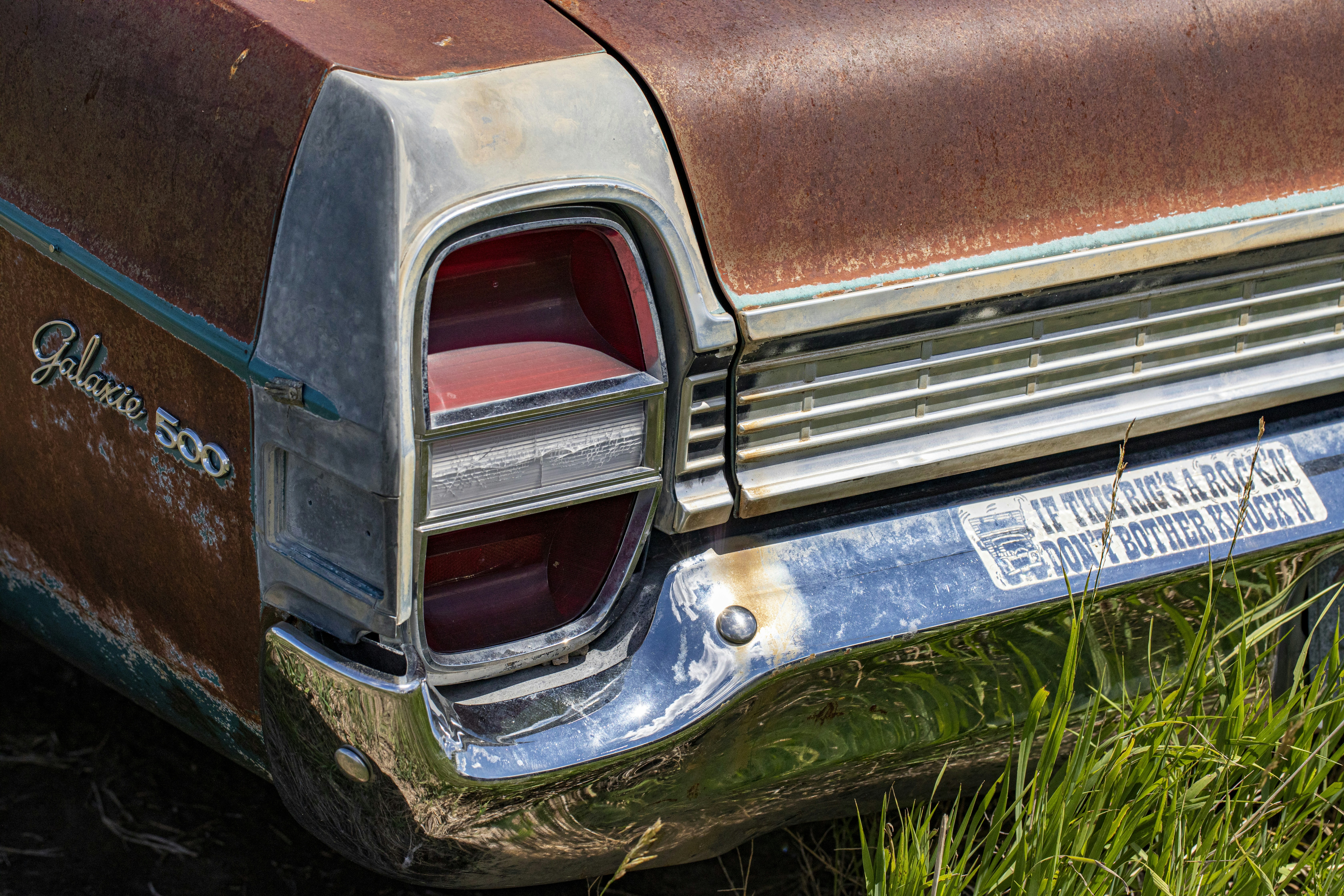 Weathered rear of a vintage Galaxie 500 showcasing its chrome details and faded paint. A humorous bumper sticker adds character.
