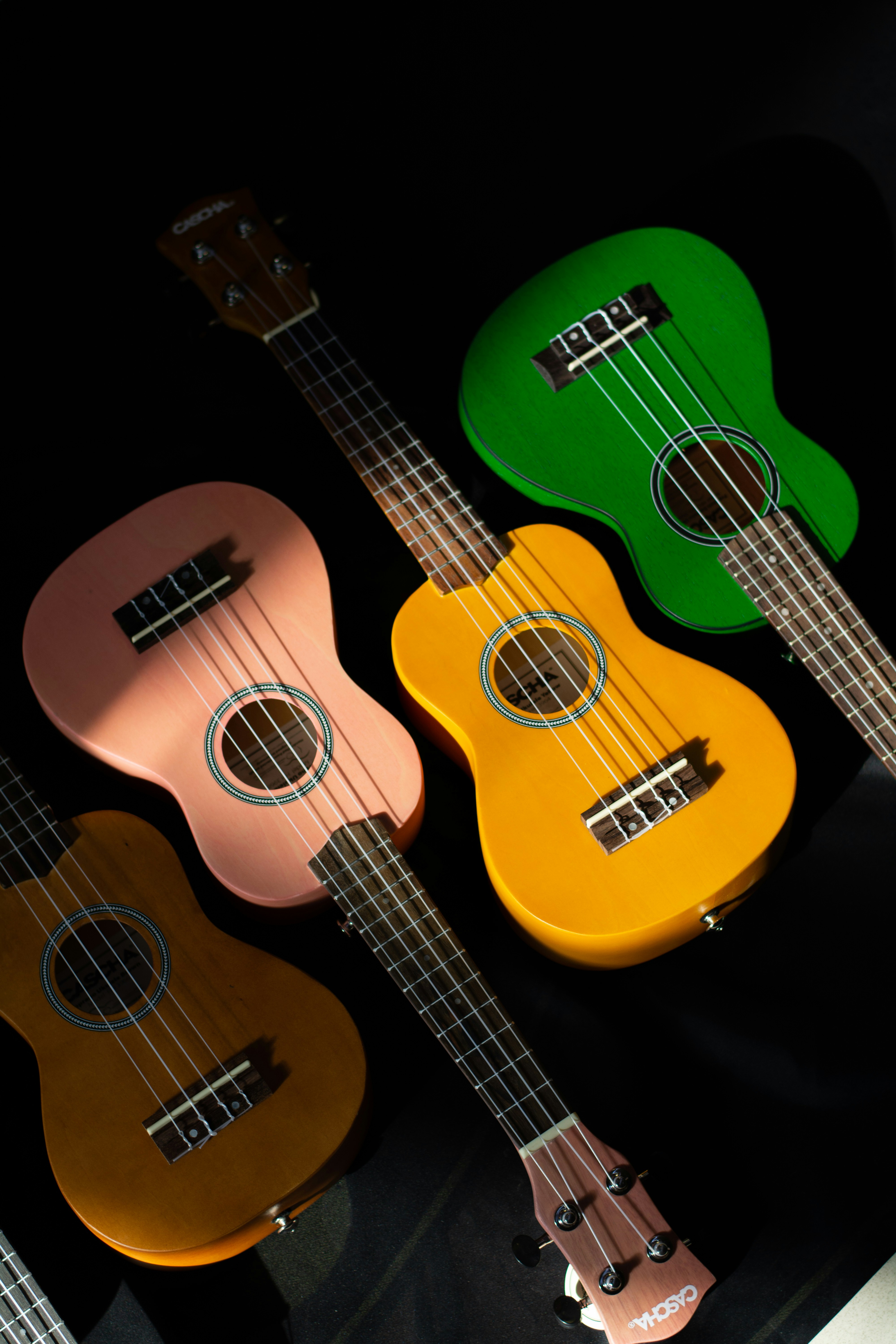 Four vibrant ukuleles in shades of pink, orange, yellow, and green arranged artistically against a dark background.