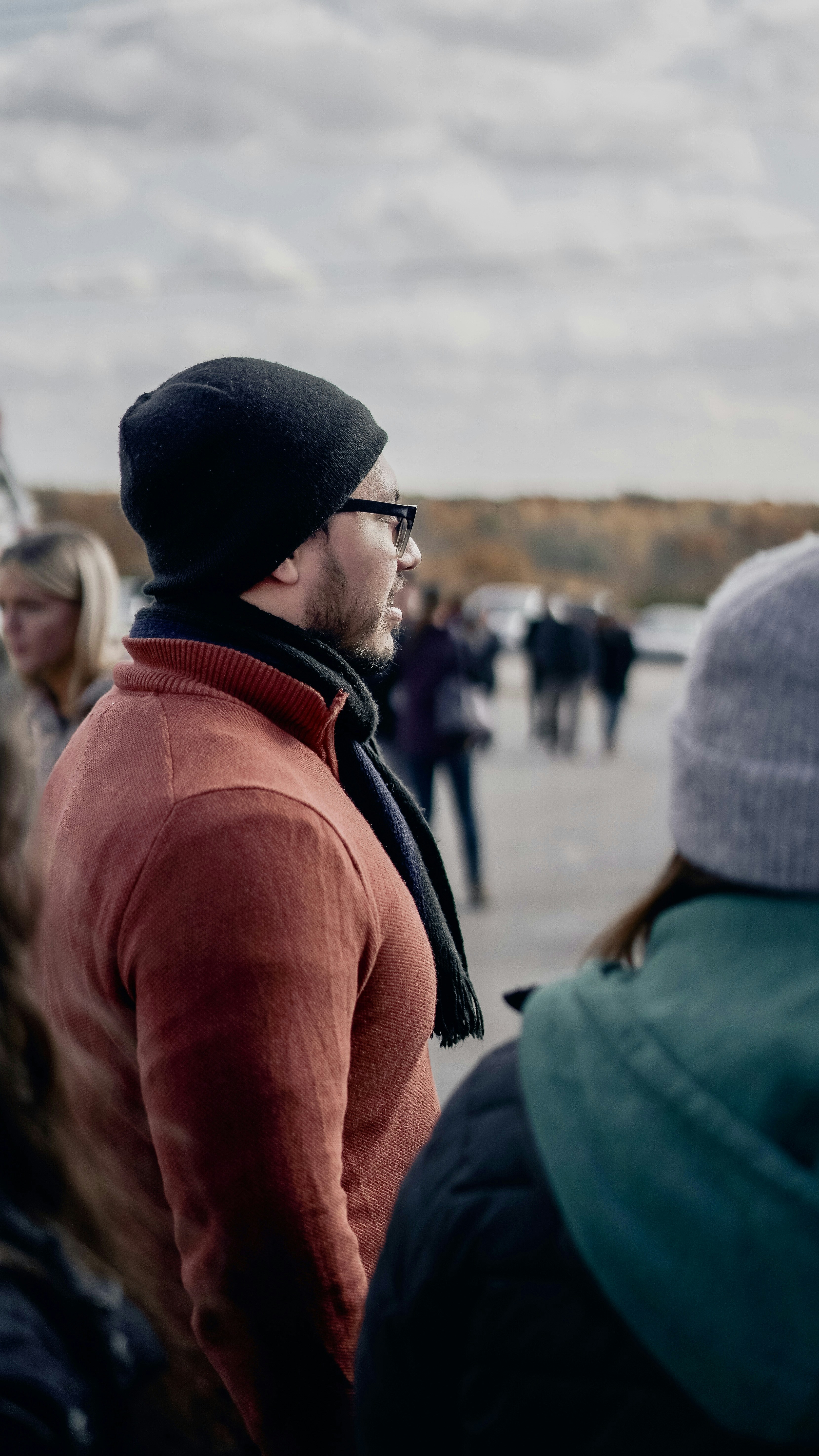 A man in a red sweater and black scarf stands thoughtfully amidst a crowd, with a blurred background of people and a cloudy sky.