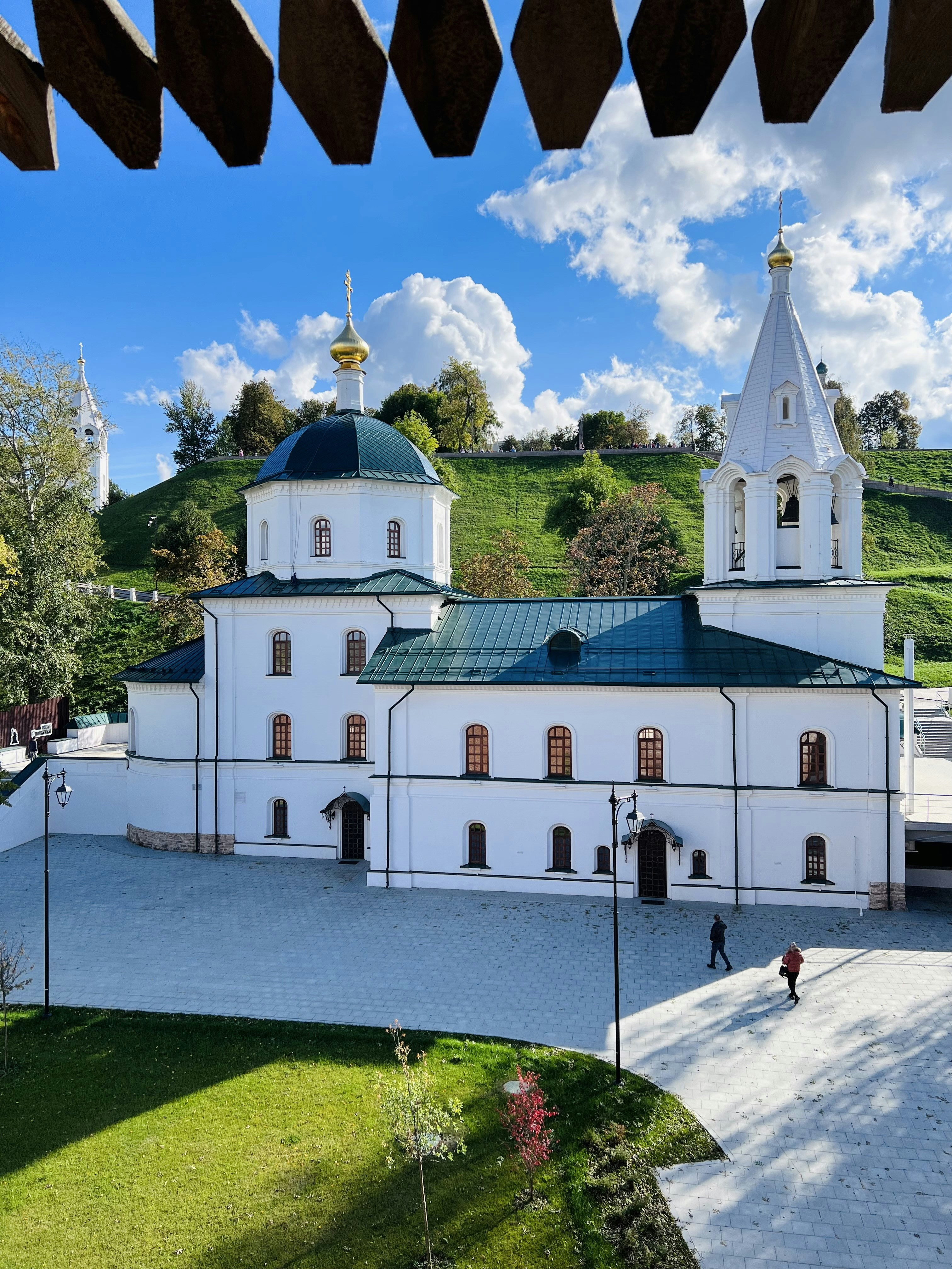 Historic church complex with a green hill backdrop and people strolling on a stone pathway.