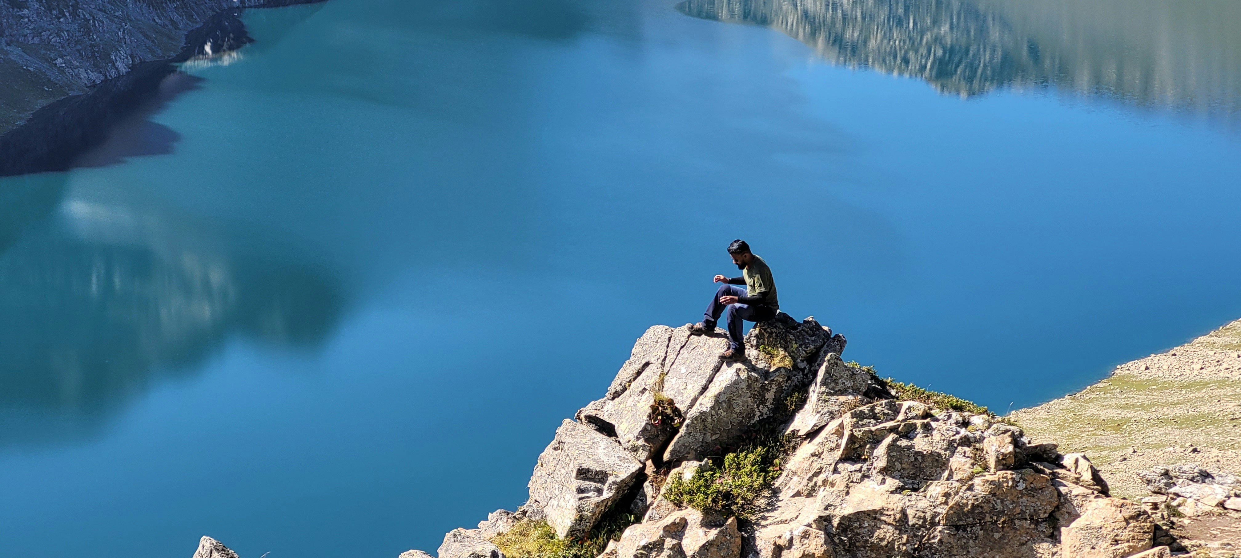 Person sitting on a rocky outcrop overlooking a tranquil blue lake with distant mountains.