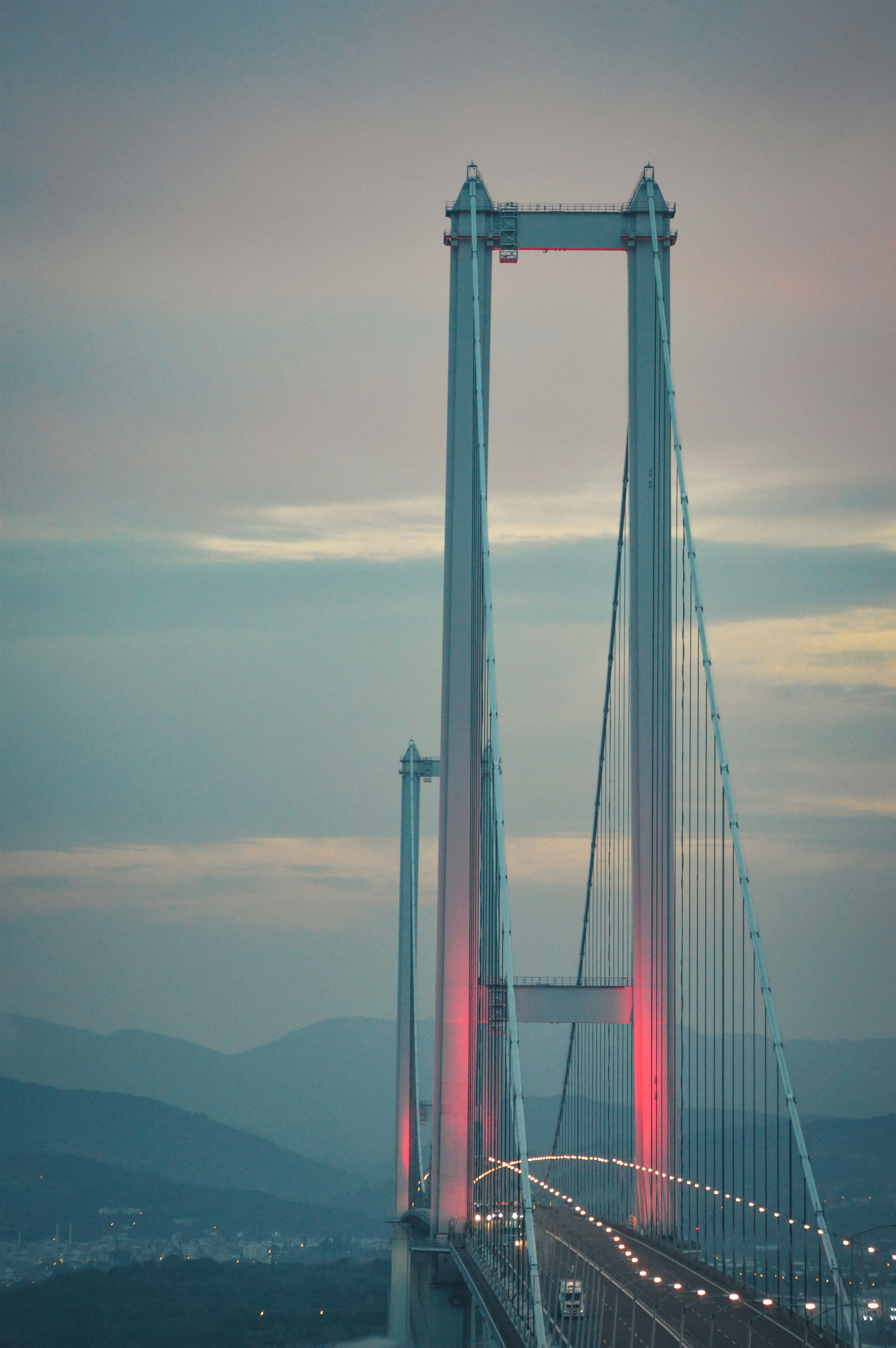 Tall suspension bridge with glowing red lights against a dusky sky.