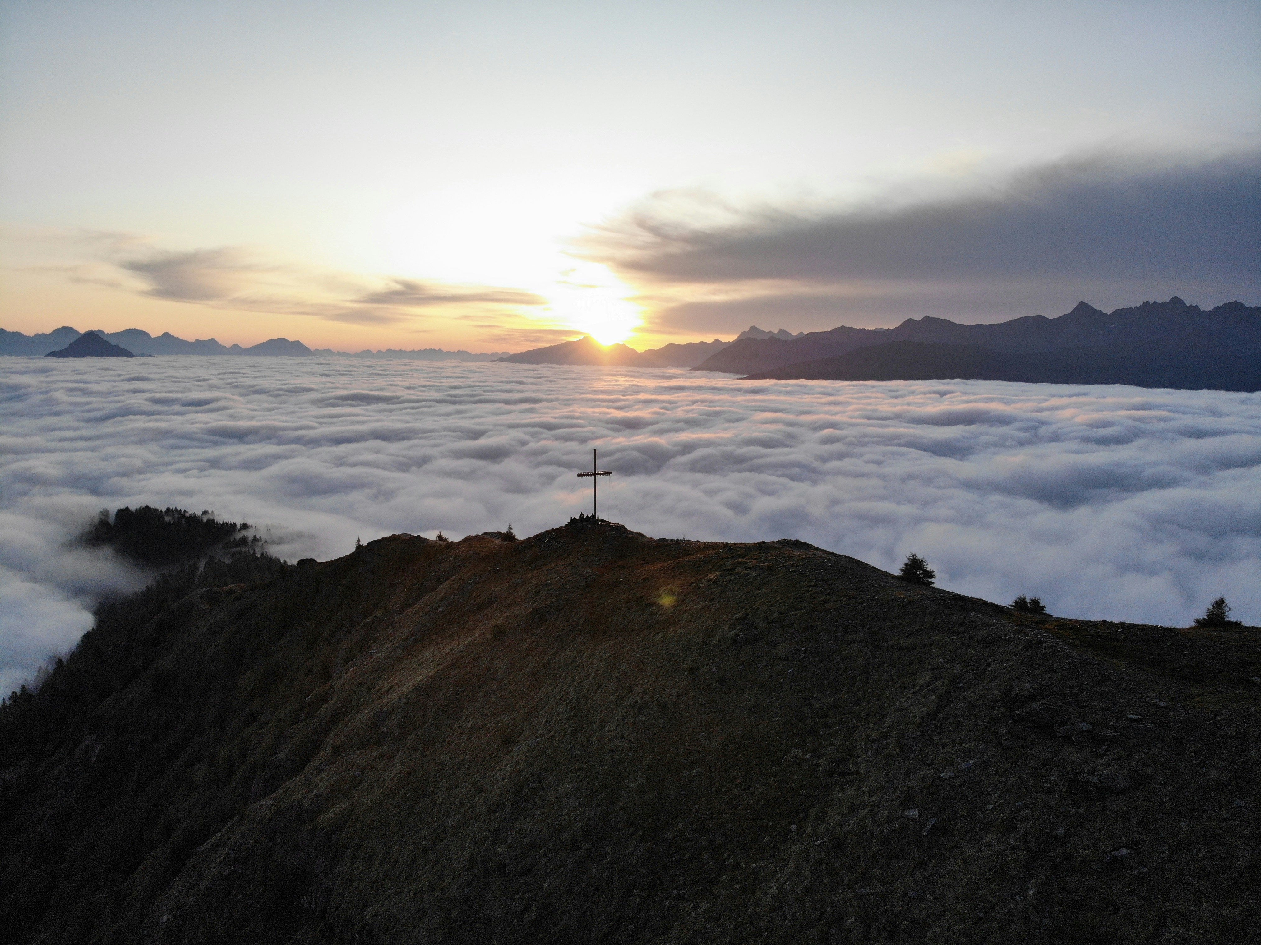 Mountain ridge with a cross silhouetted against a sea of clouds and the setting sun.