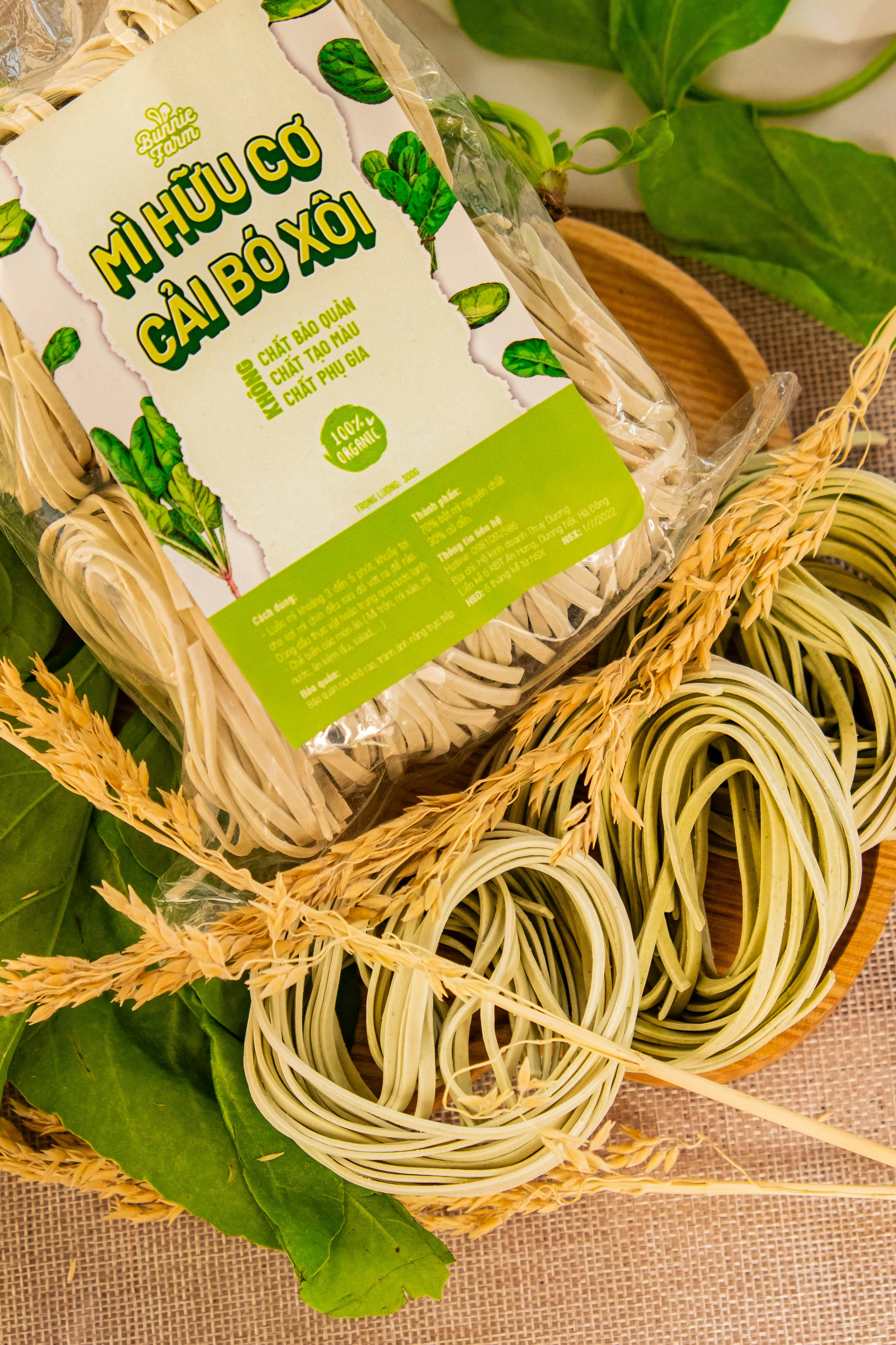 Packaged organic noodles accompanied by fresh greens and wheat stalks, showcasing a blend of natural ingredients.