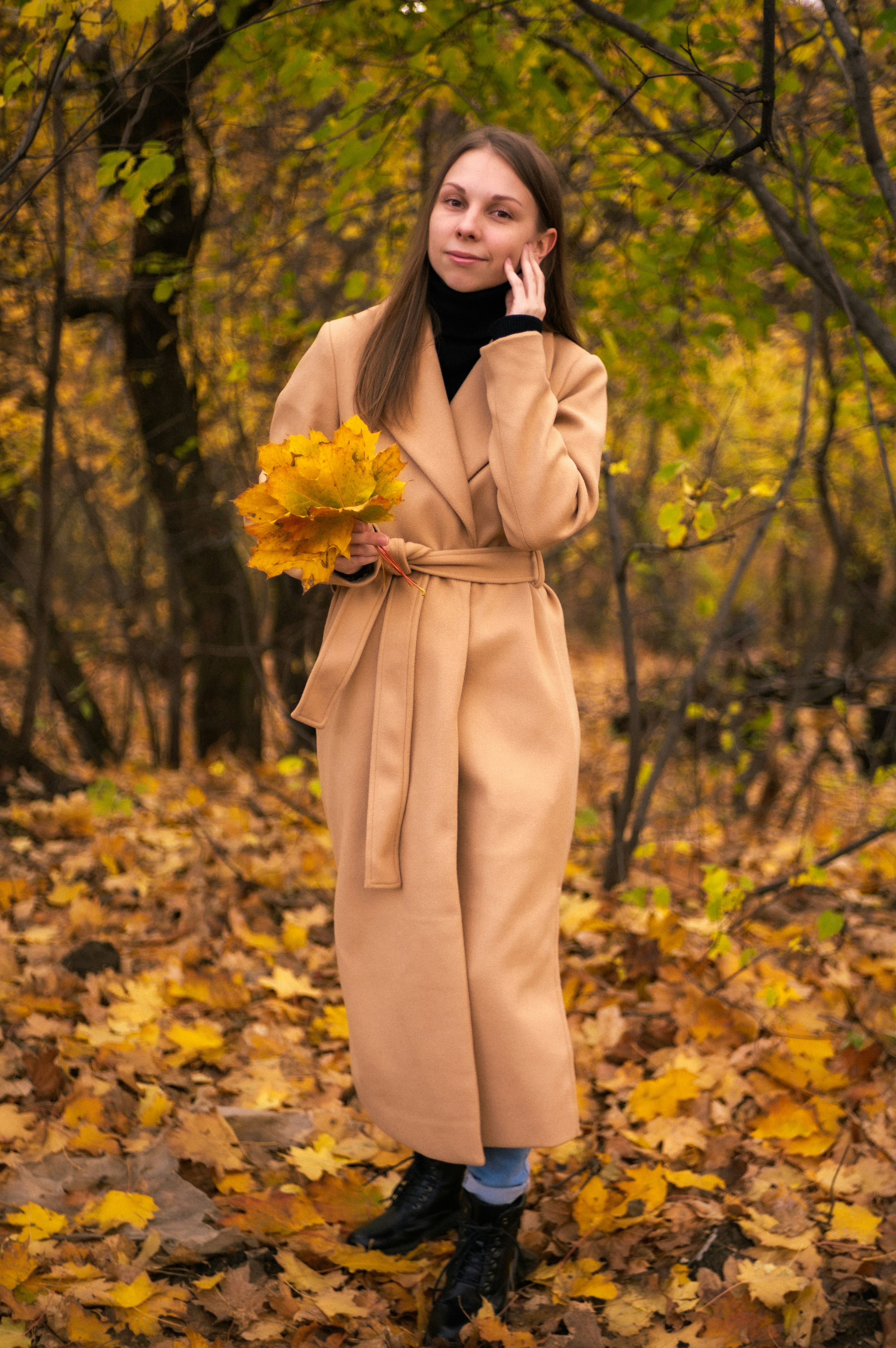 Autumn forest portrait of a woman in a camel coat holding a bouquet of yellow autumn leaves.
