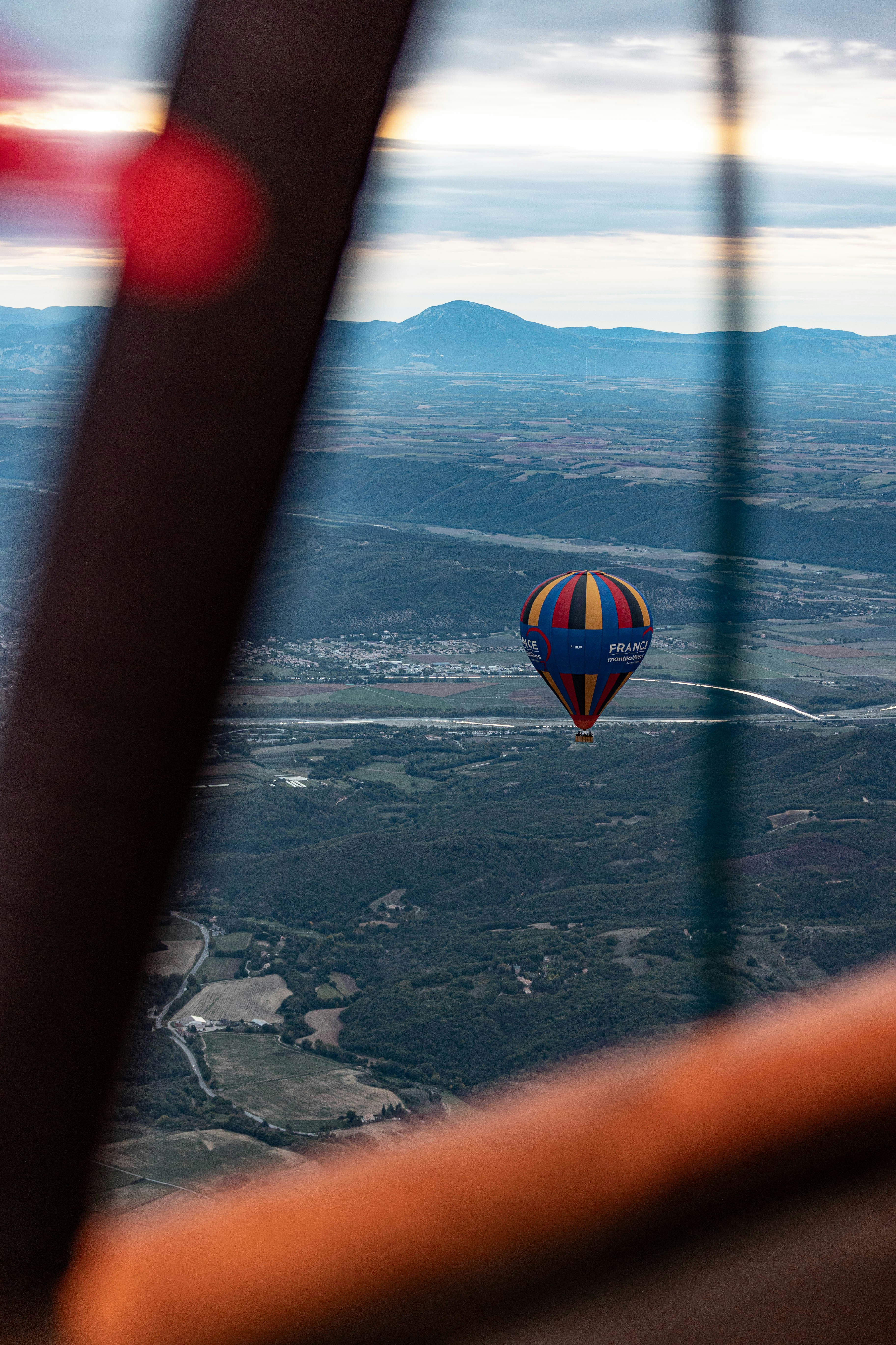 Stripes Over the Valley: A distant hot air balloon glides above patchwork fields framed by the basket’s foreground ribs