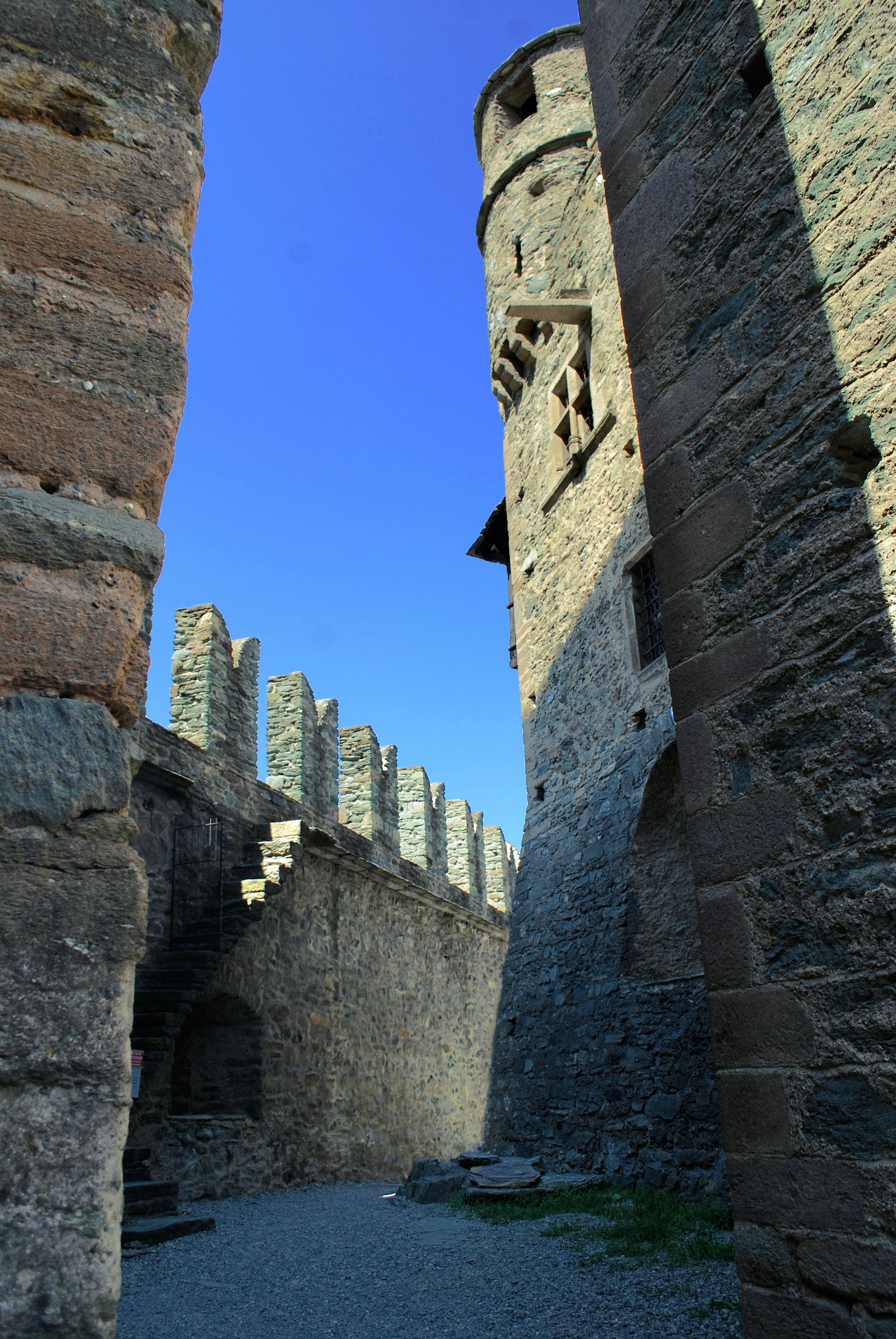 Weathered stone walls and a staircase lead through a historic castle courtyard, framed by towering battlements against a clear blue sky.