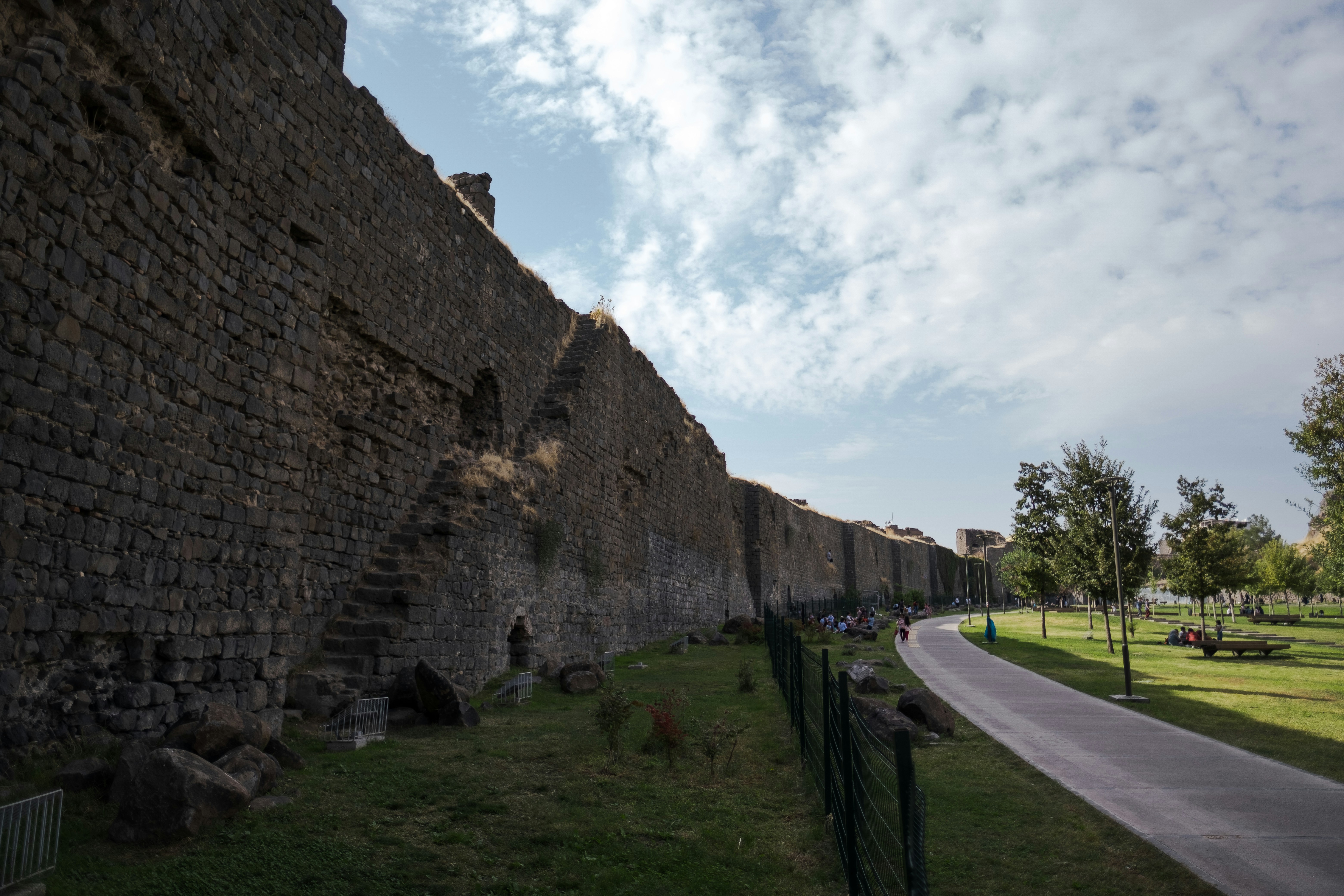 Historic stone wall stretches alongside a park, with visitors enjoying the greenery and pathways nearby.