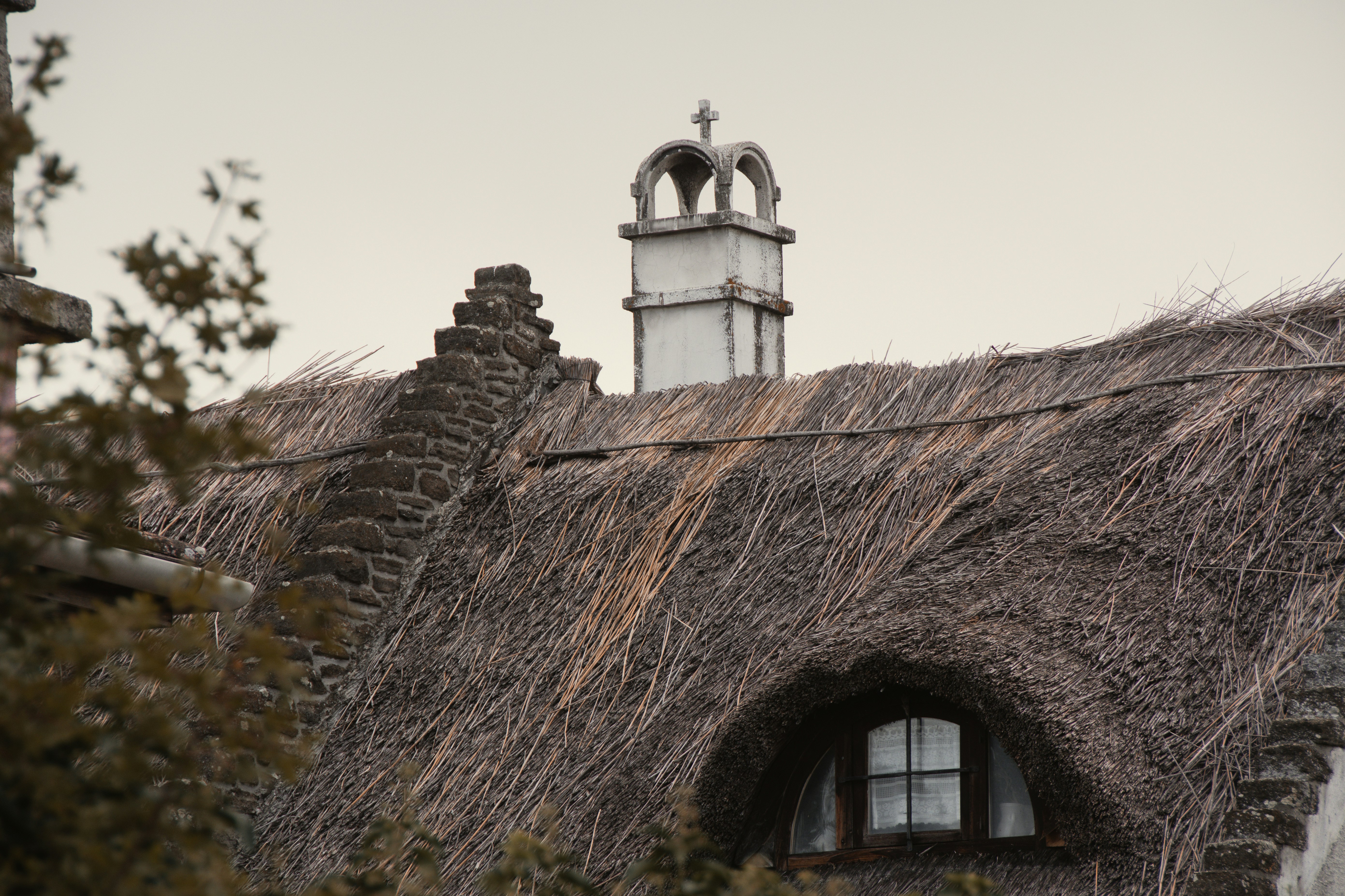 A rustic thatched roof with a prominent chimney and window, showcasing traditional architecture against a muted sky.