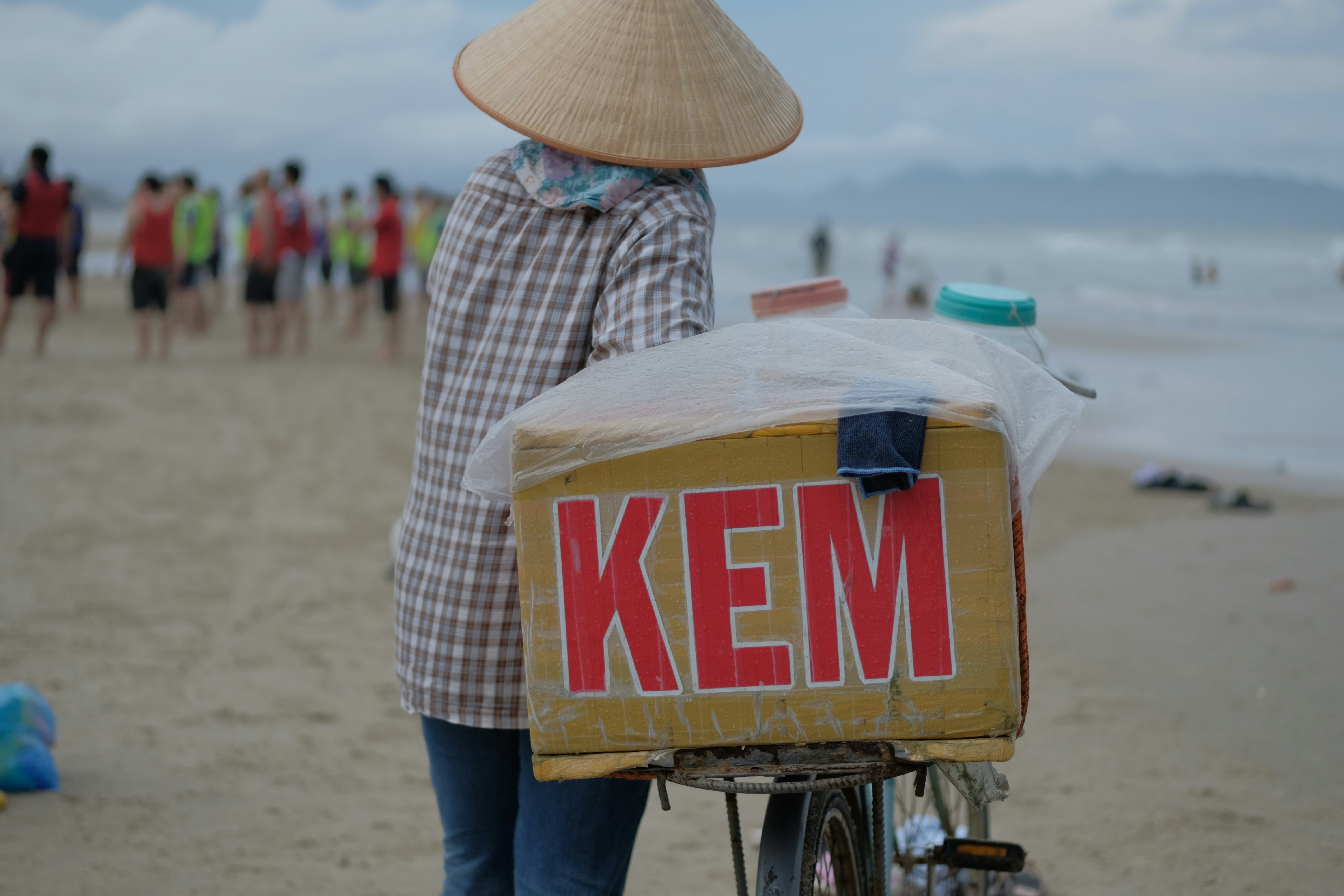 Ice cream vendor with conical hat pushes cart along a busy beachside path.