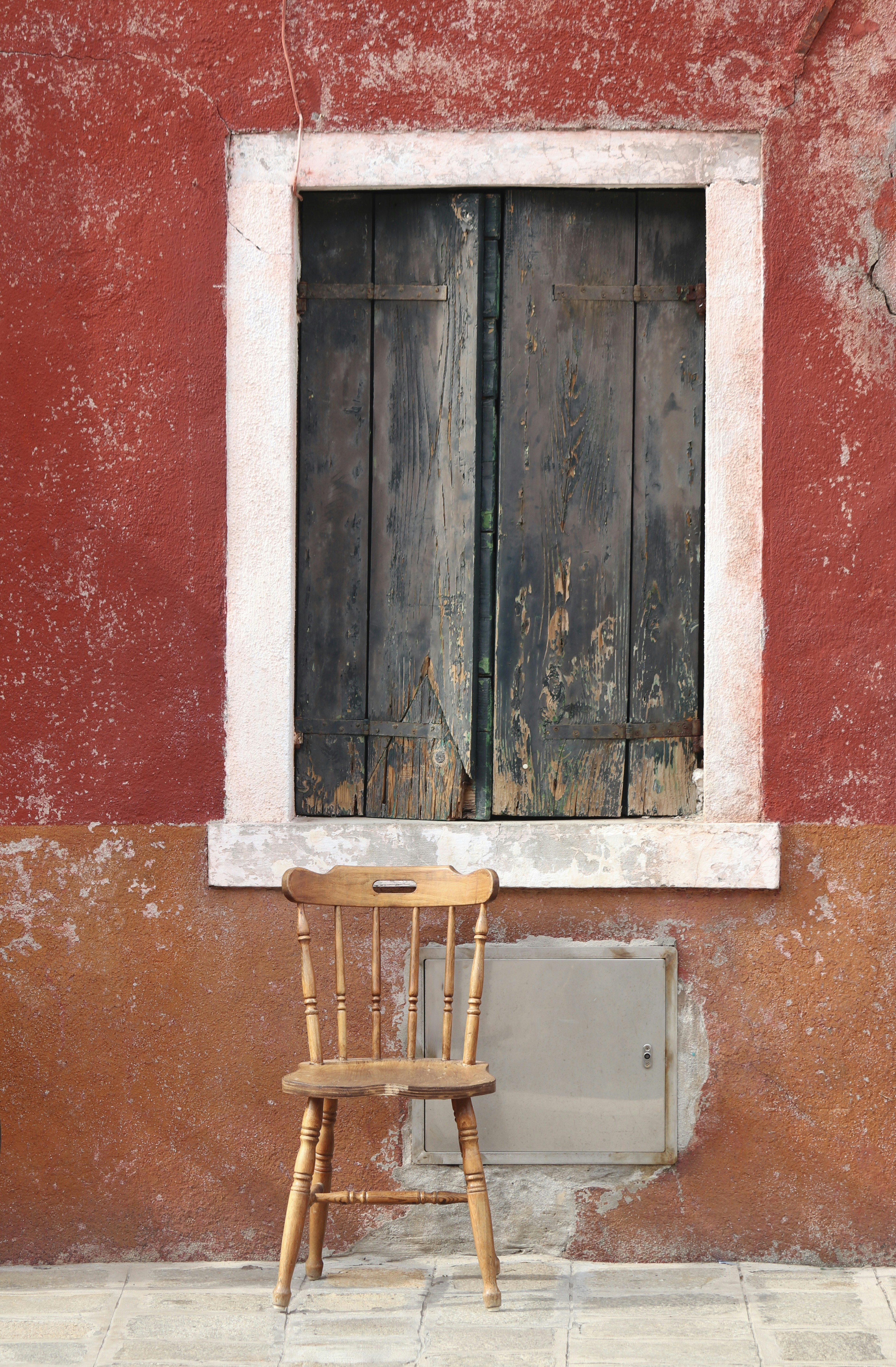 A rustic wooden chair sits in front of a weathered window with dark shutters against a textured red wall. The scene evokes a sense of nostalgia and quiet reflection.