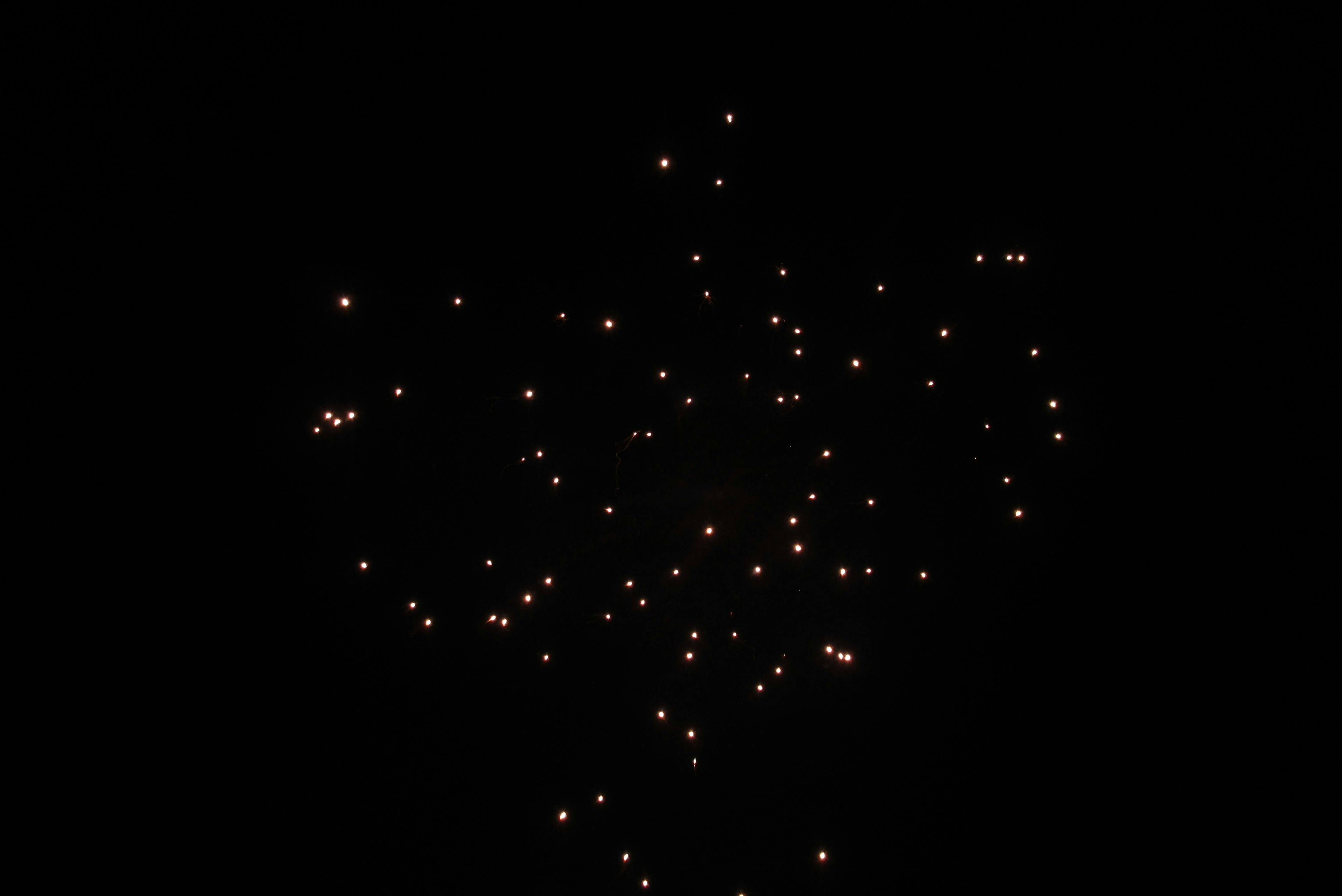 Cluster of small orange sparks scattered across a dark sky resembles a sparse fireworks display. The shot foregrounds delicate ember highlights against deep shadows.