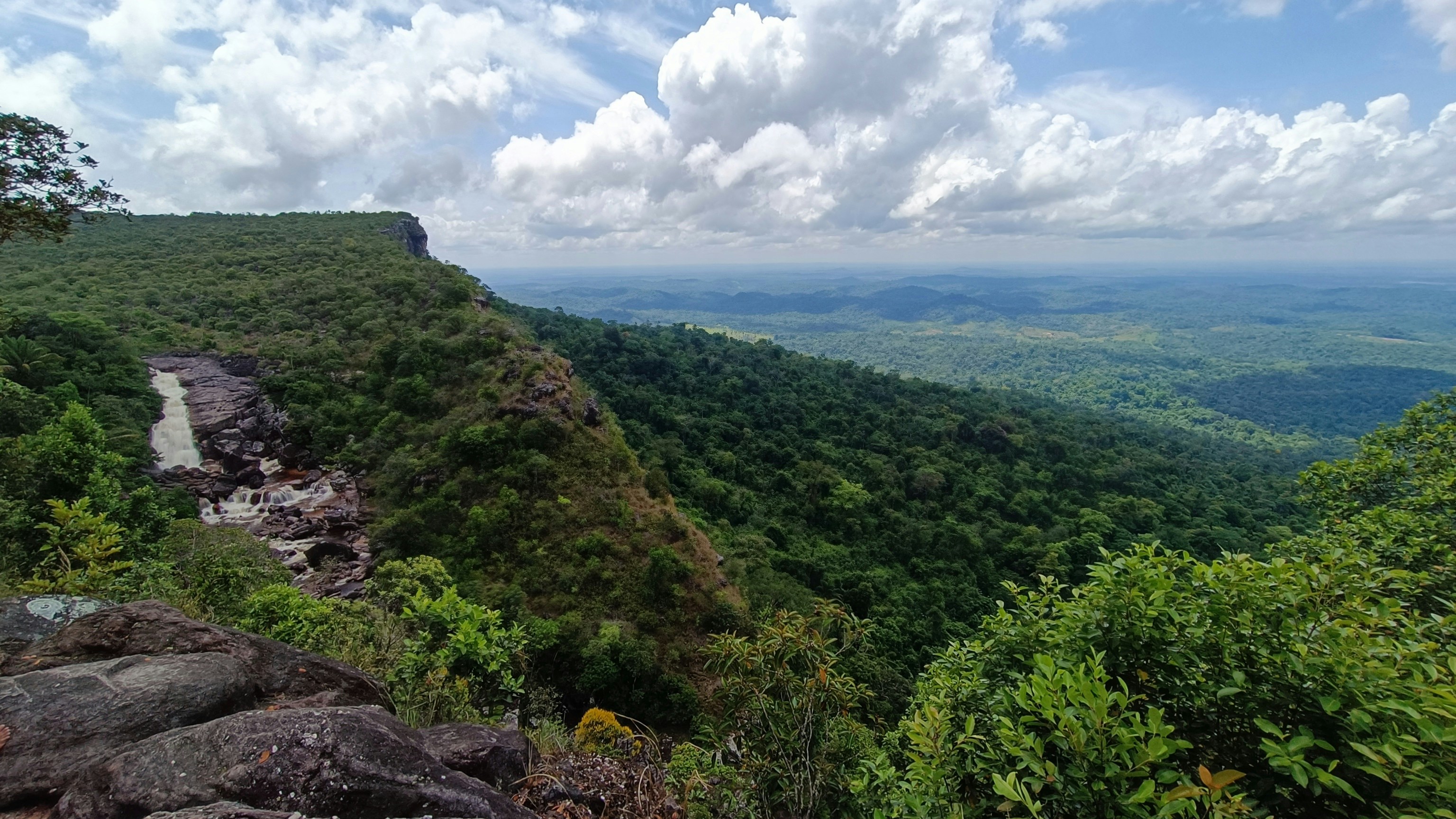 Expansive view of a lush, green valley bordered by rocky cliffs under a sky filled with billowing clouds.