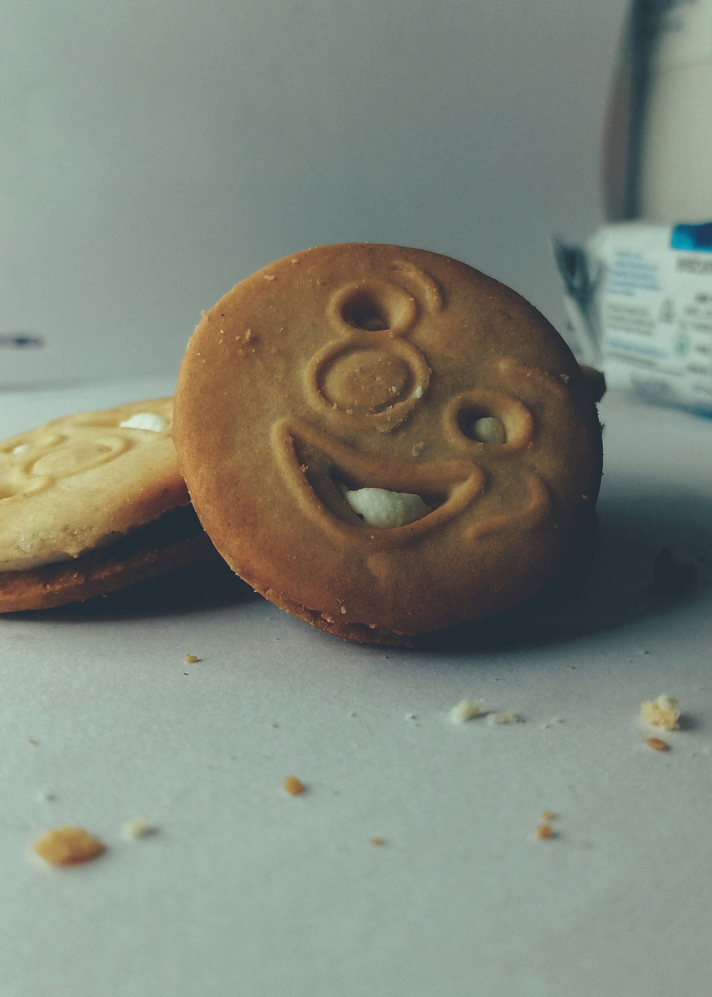 A smiling round biscuit sits on a pale surface with scattered crumbs, a second cookie softly blurred in the background.