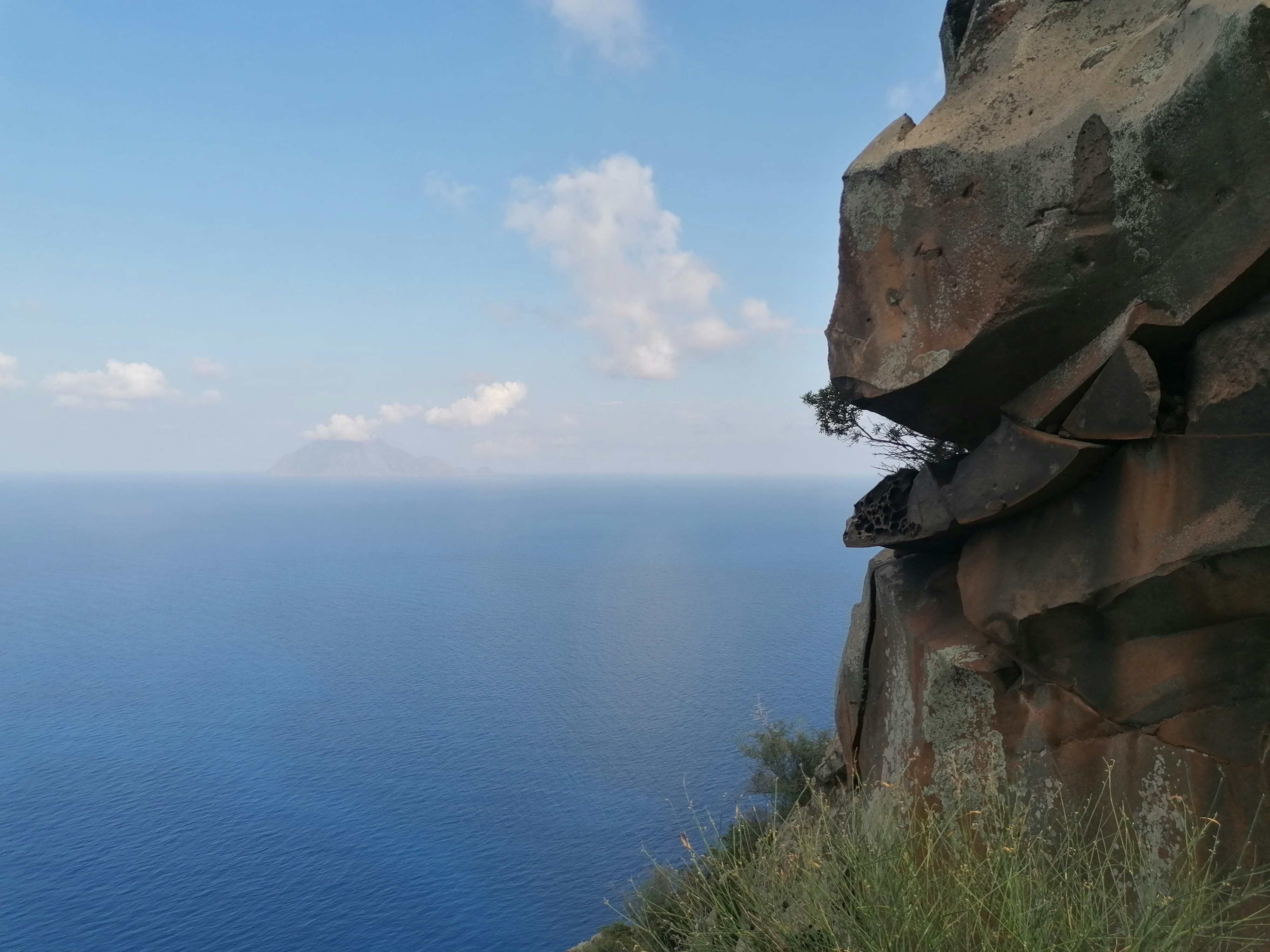 Jagged cliff edge with sparse vegetation against a vast blue sea and distant islands under a clear sky.
