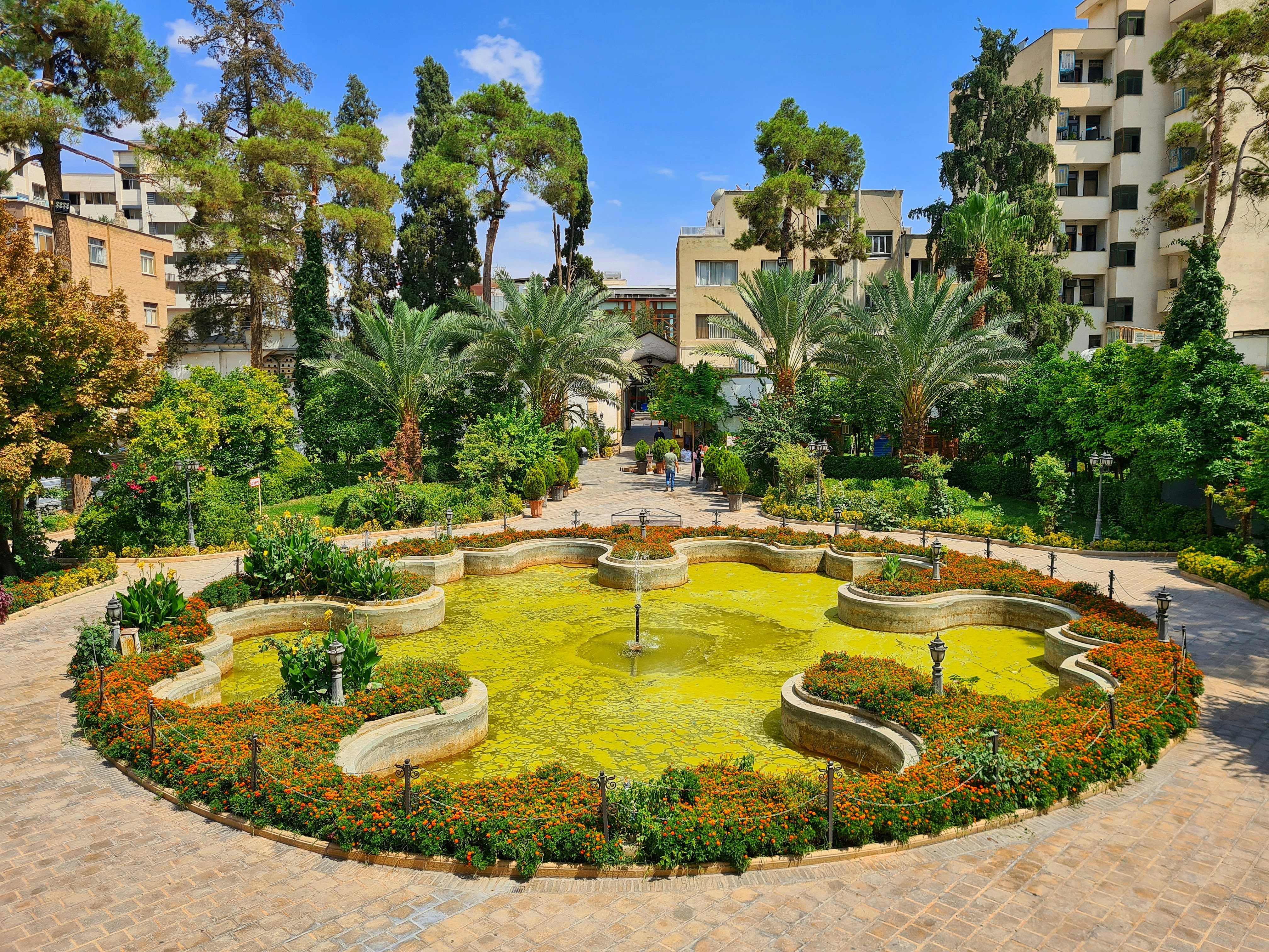 Lush greenery and a vibrant fountain surrounded by urban architecture under a clear blue sky.