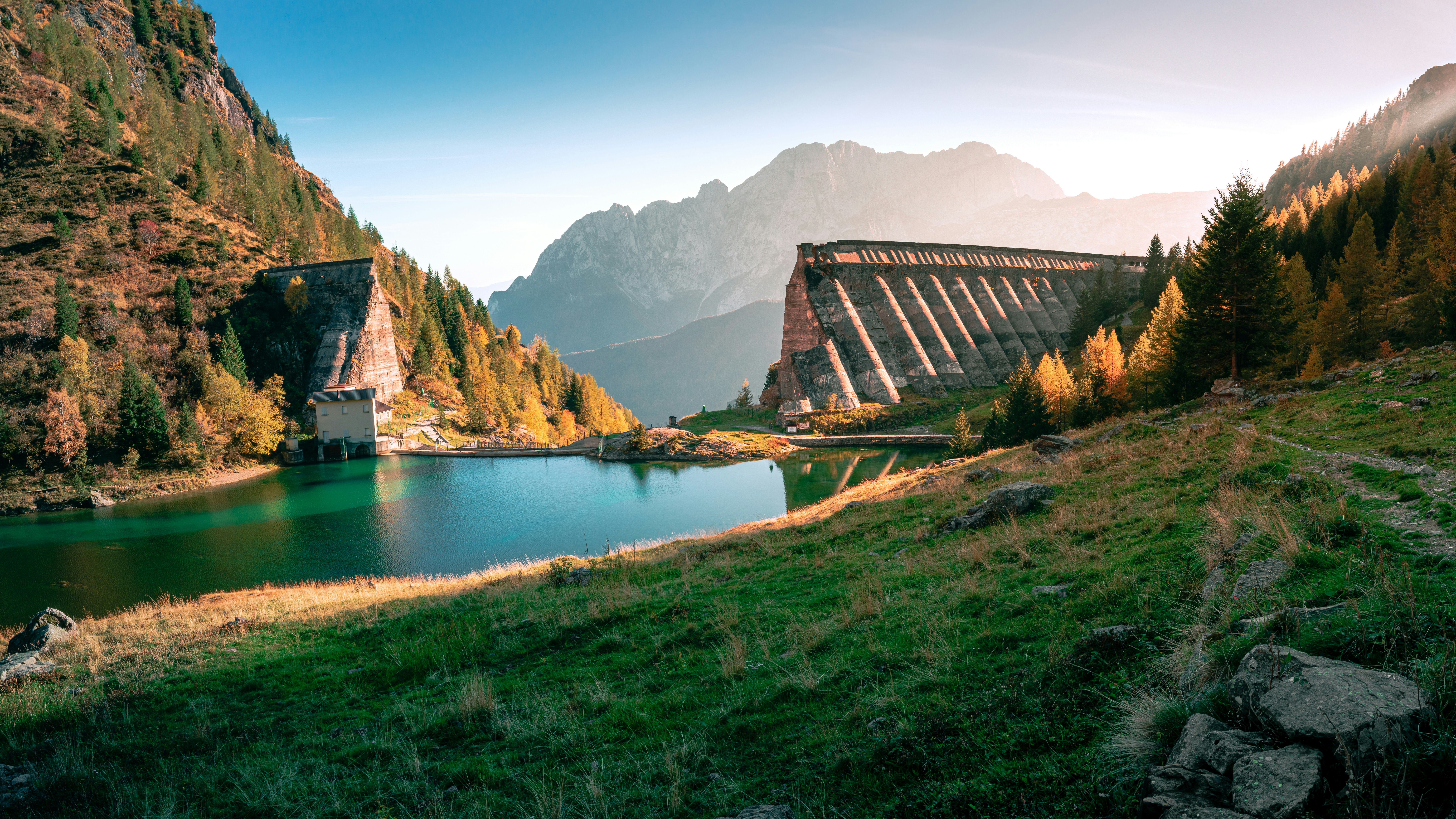 A dam rests alongside a tranquil lake surrounded by autumnal forests and distant mountains.